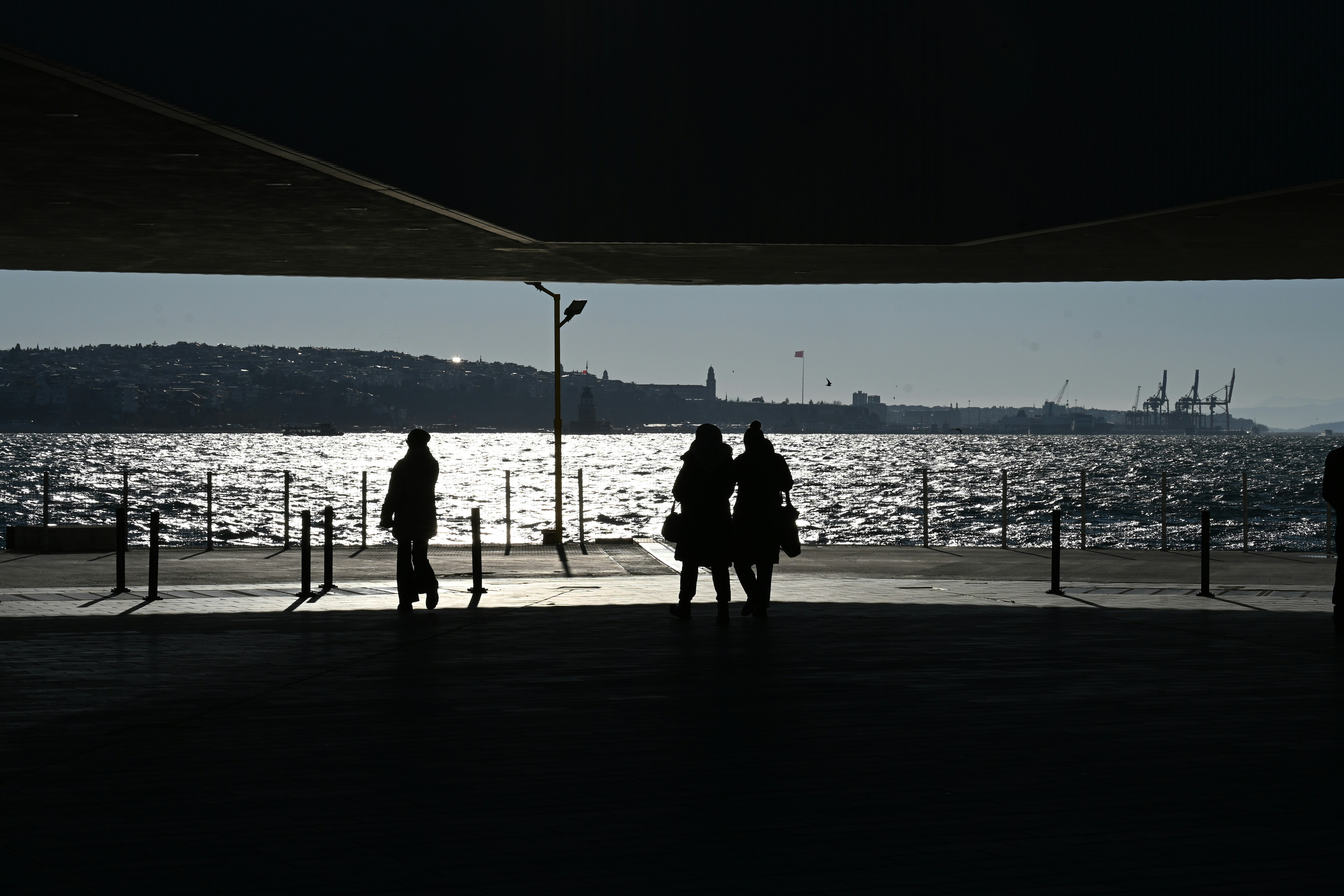 Silhouetted figures beneath a shaded overhang overlooking a sunlit harbor