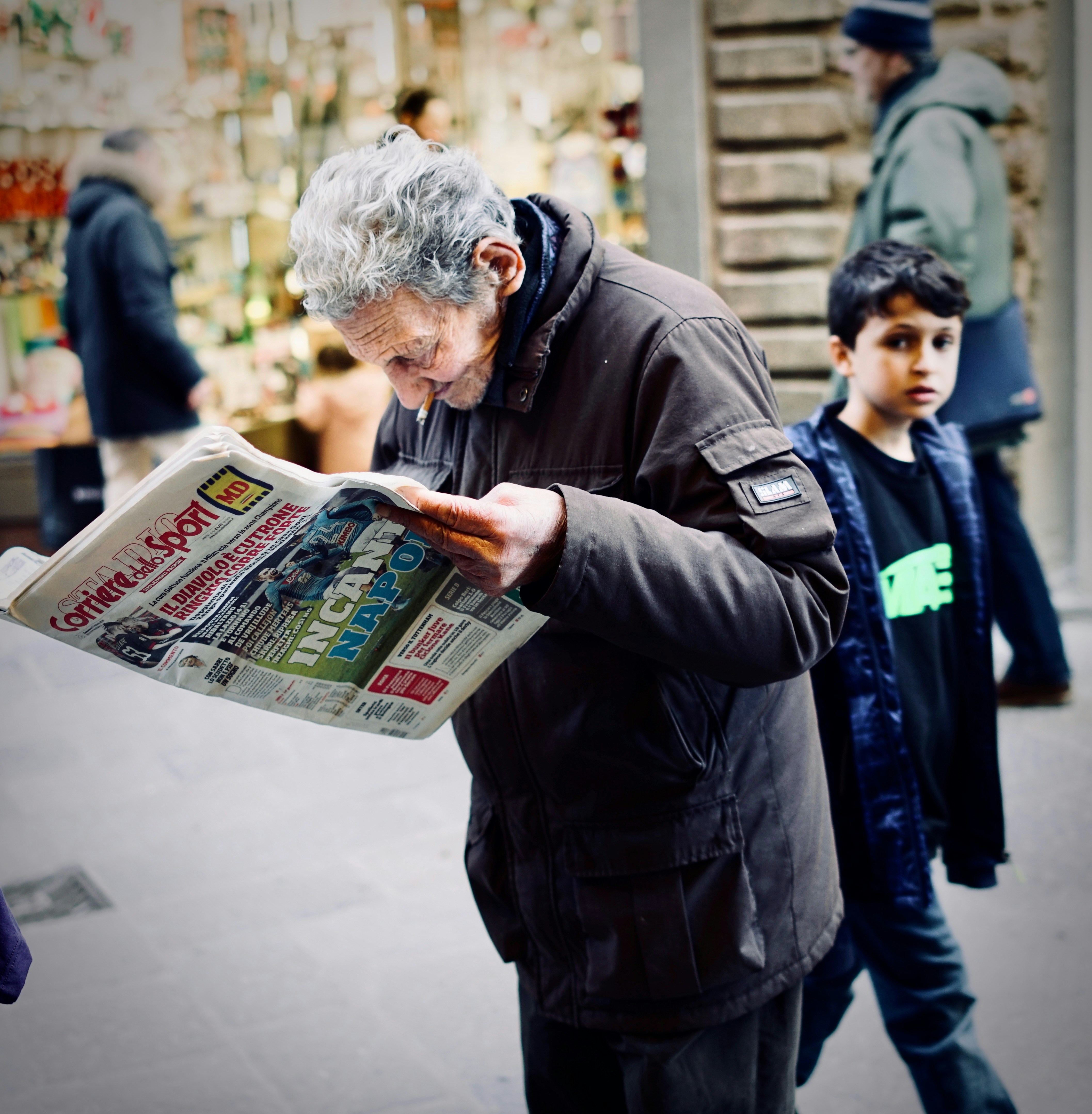Elderly man reads newspaper while boy stands nearby.