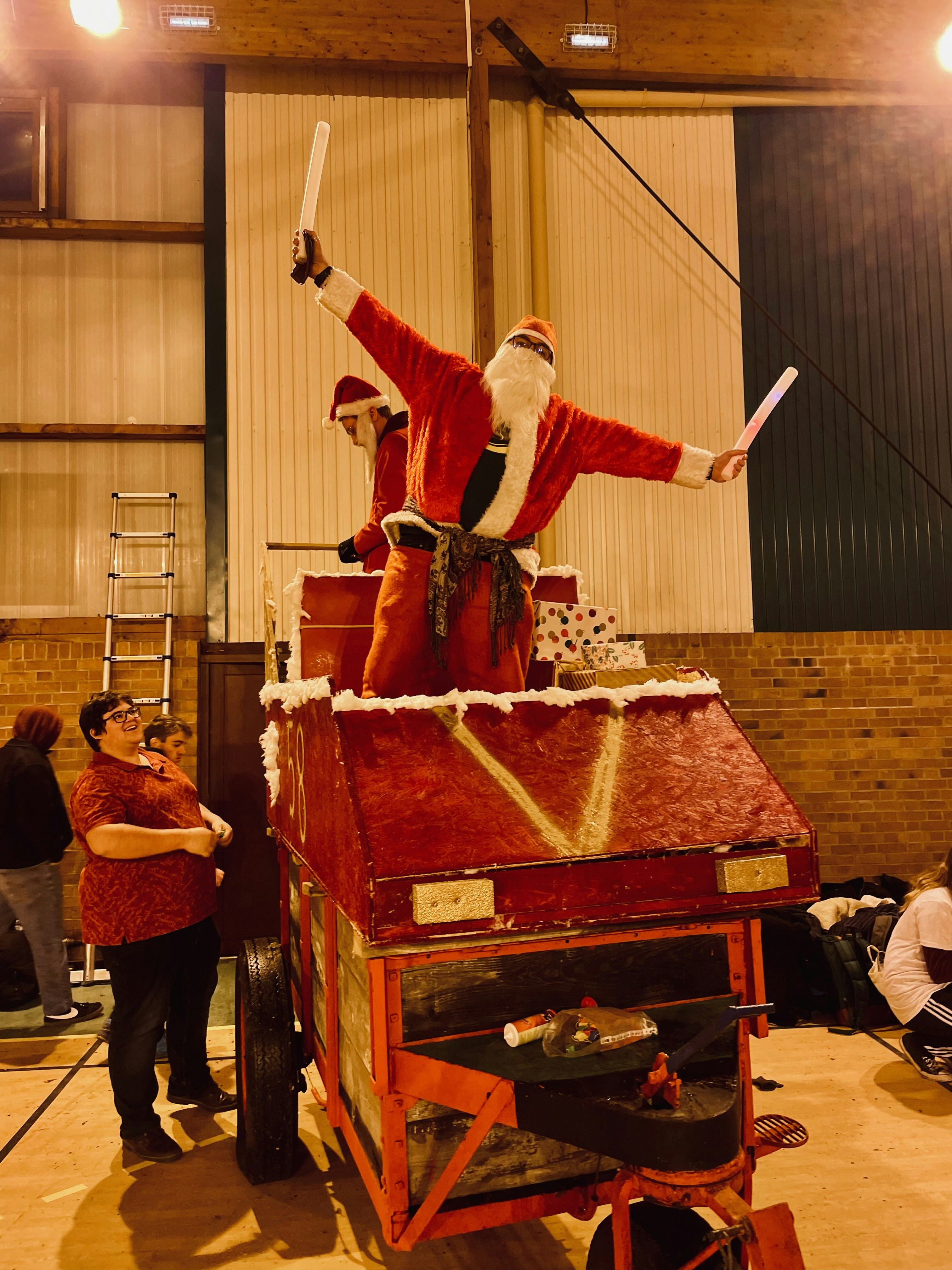 Brightly lit indoor scene with Santa Claus standing atop a red decorated cart, arms raised with glow sticks, while helpers watch nearby in a gym-like hall.