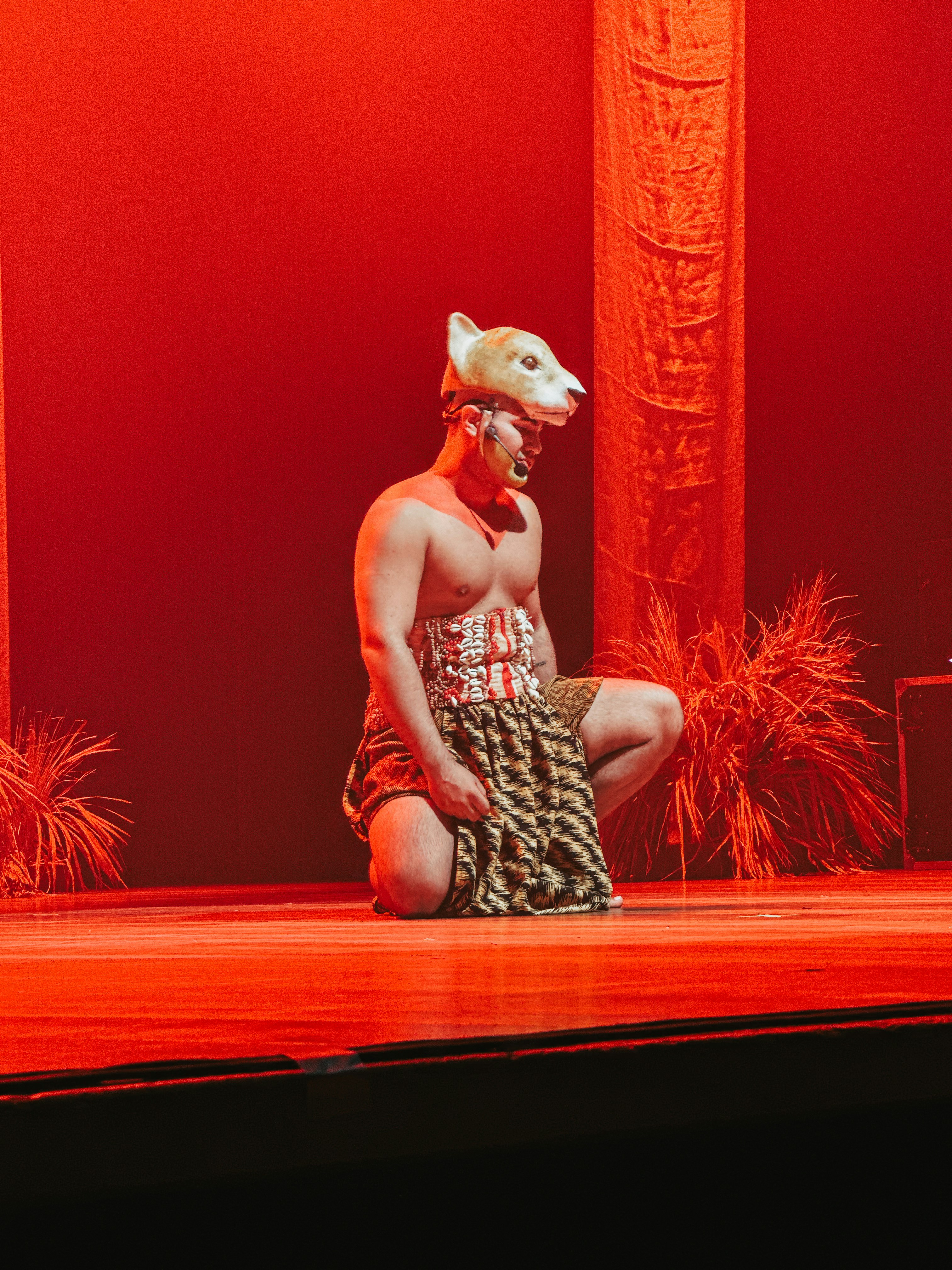 Masked performer on a red-lit stage kneels beside a tall pillar