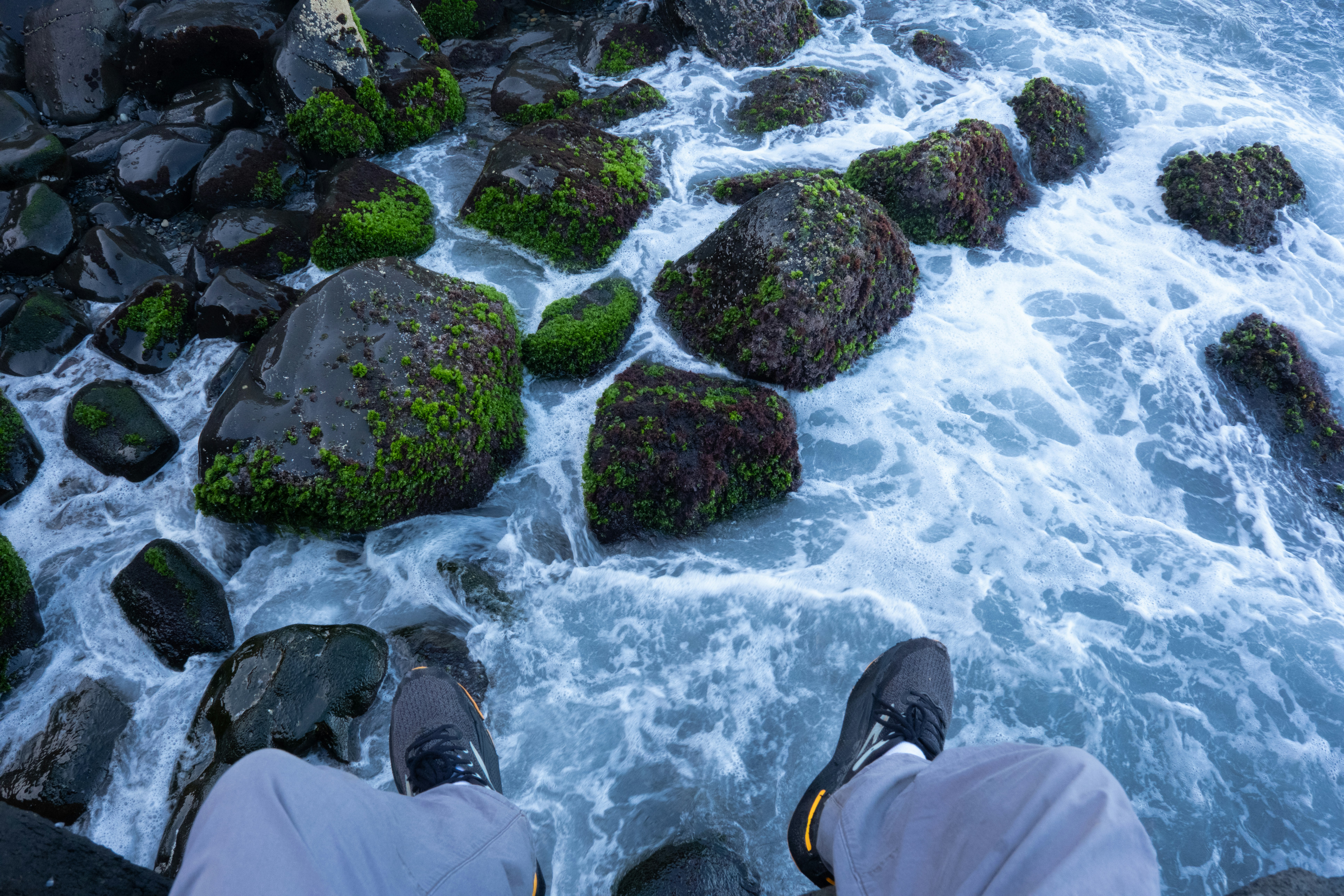 Over the Edge: Boots on Mossy Rim Over Turbulent Tides