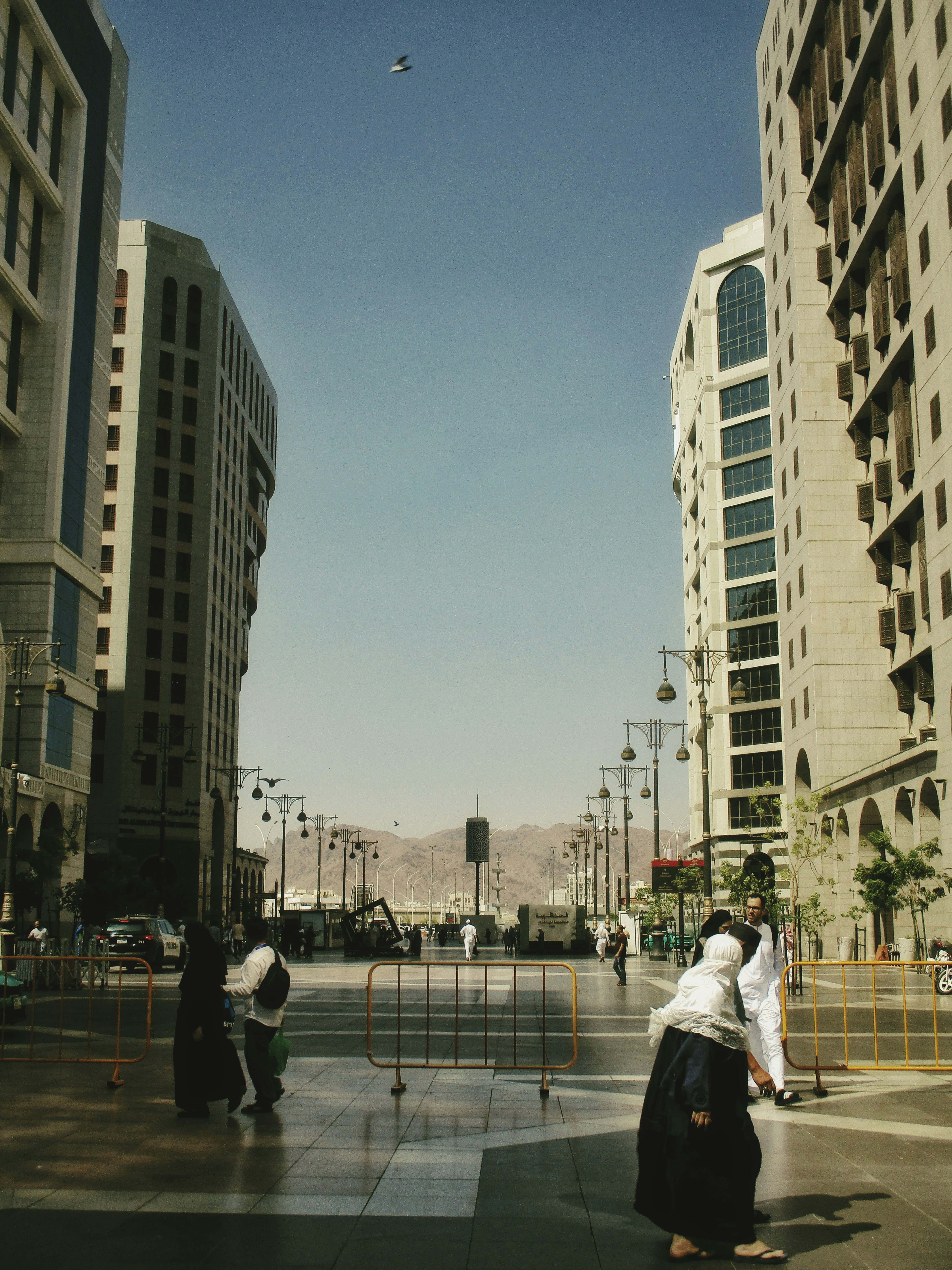 People moving through a modern urban space, surrounded by towering buildings under the Madinah sky.