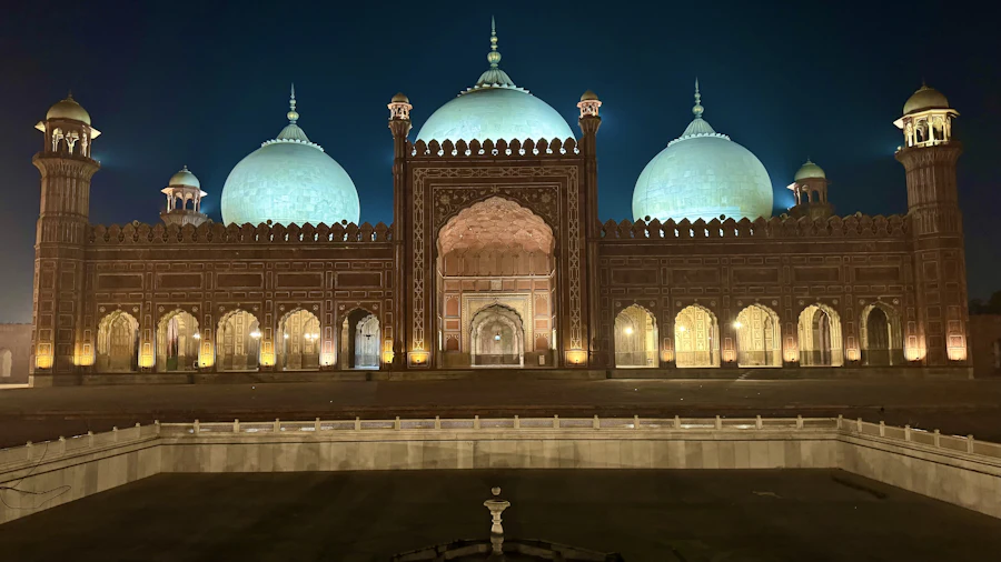 Illuminated mosque with teal domes and arched colonnade at night, reflecting golden lighting on a tranquil foreground plaza.
