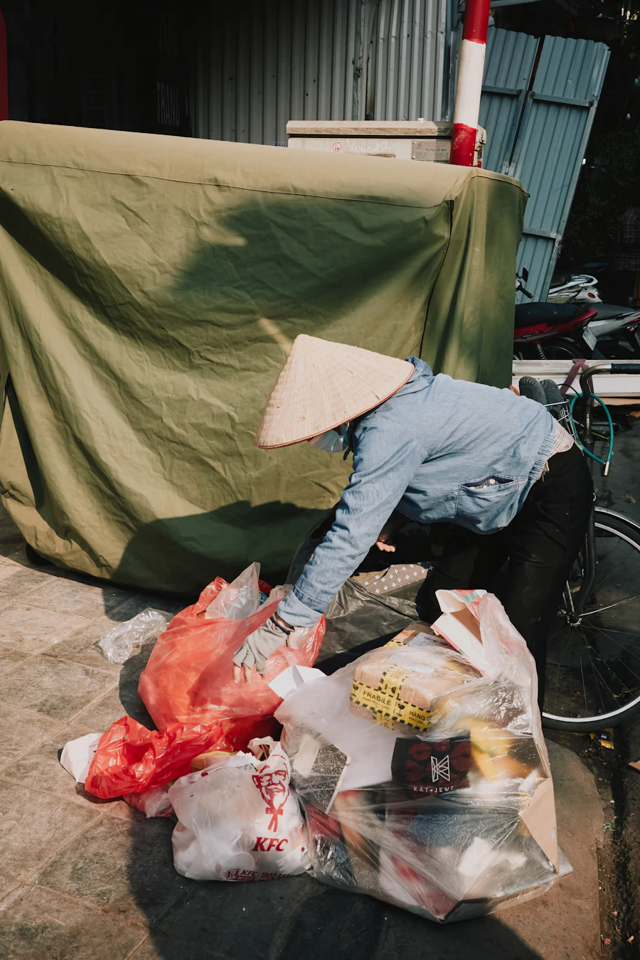 A person wearing a conical hat and denim jacket sifts through plastic bags on a sidewalk beside a parked bicycle and a corrugated metal structure.