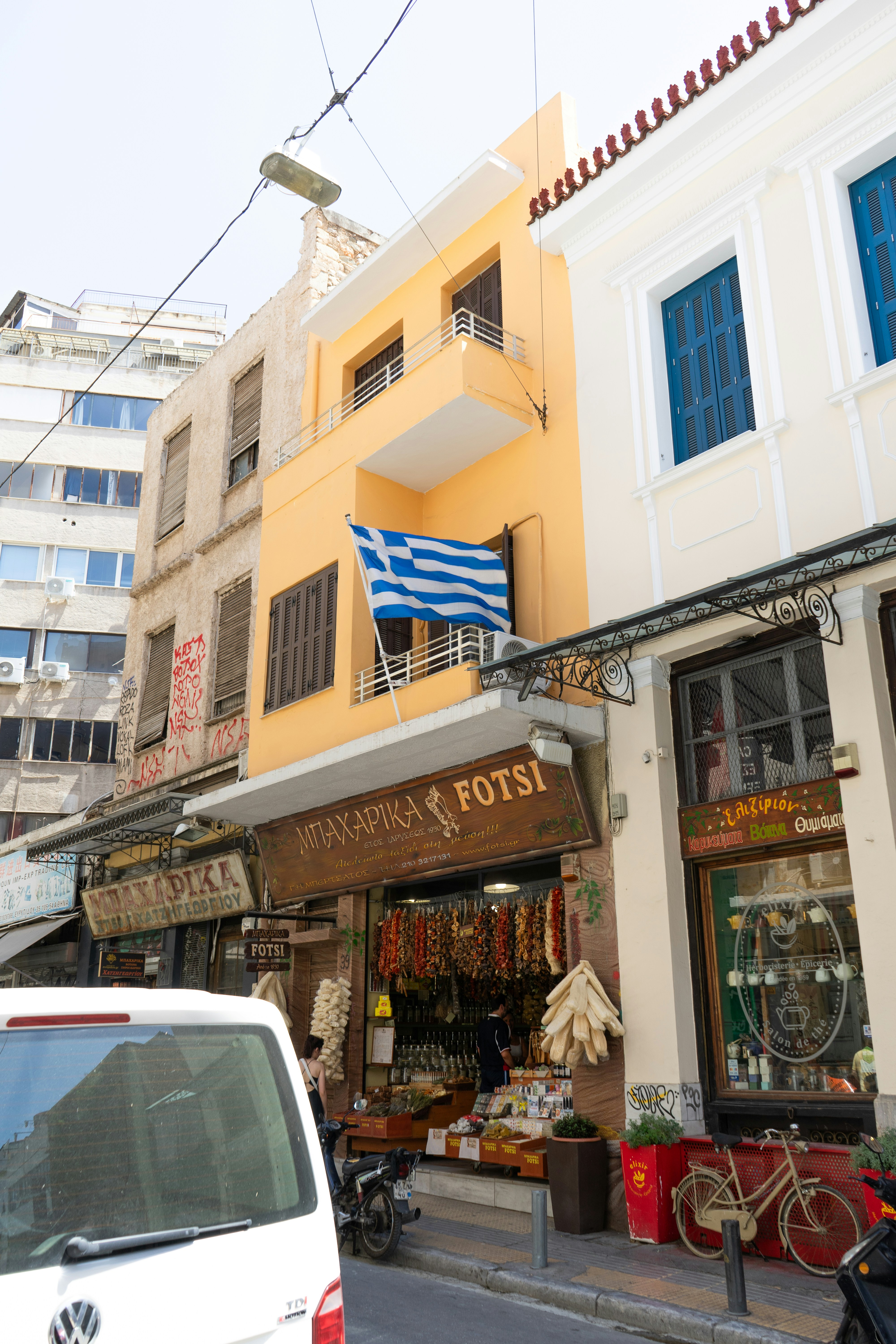 Yellow Balcony Frontage with Greek Flag Above a Street Vendor Shop