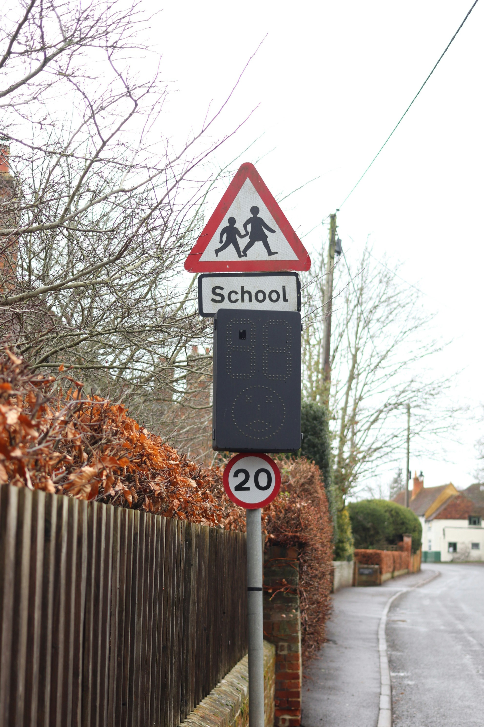 A roadside scene features a school warning sign with an electronic display and a 20 mph sign beneath, set along a narrow quiet street bordered by a wooden fence and leafless trees.