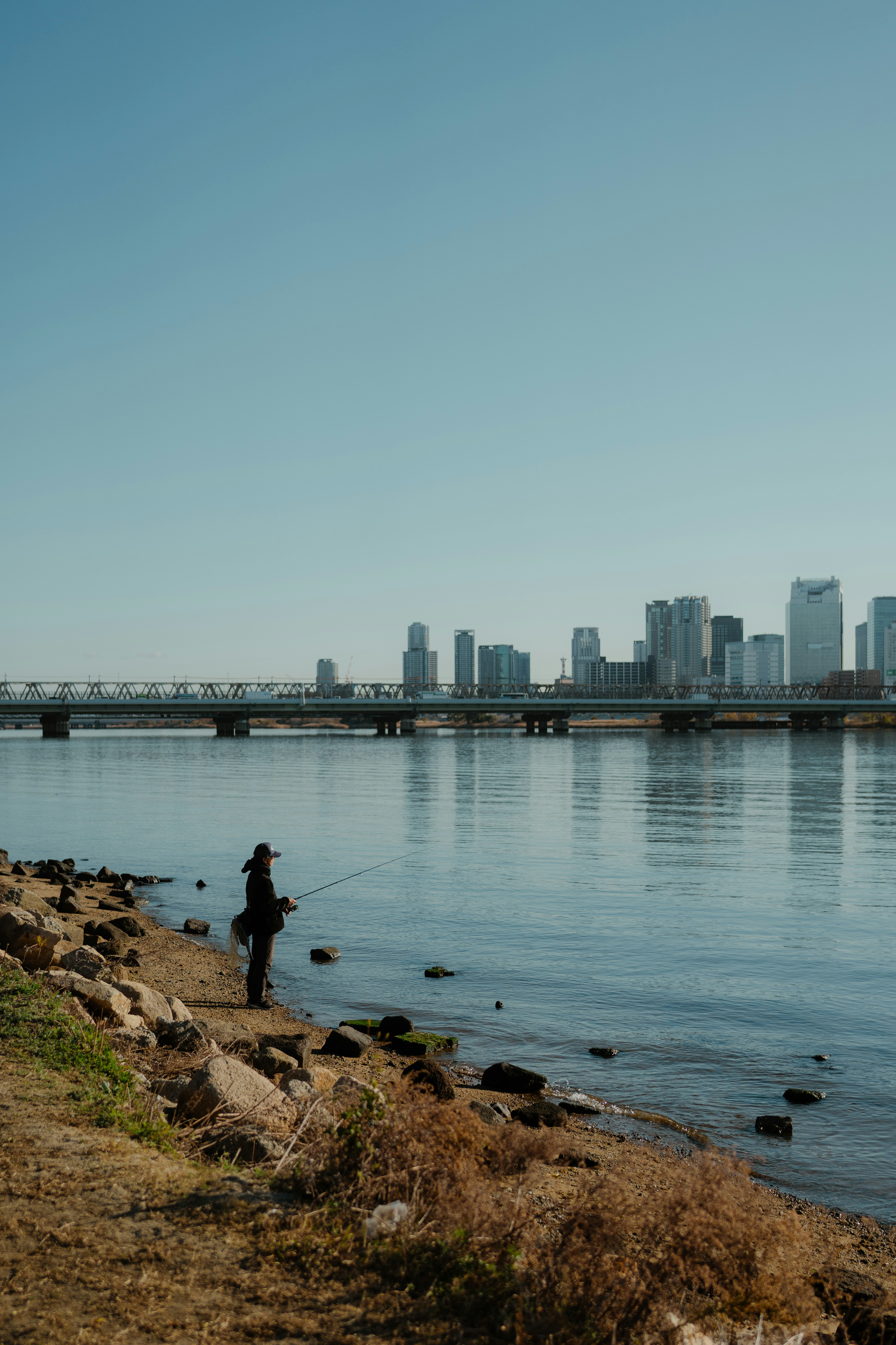 Riverside Casting: A Fisherman, City Silhouette, and Calm Waters