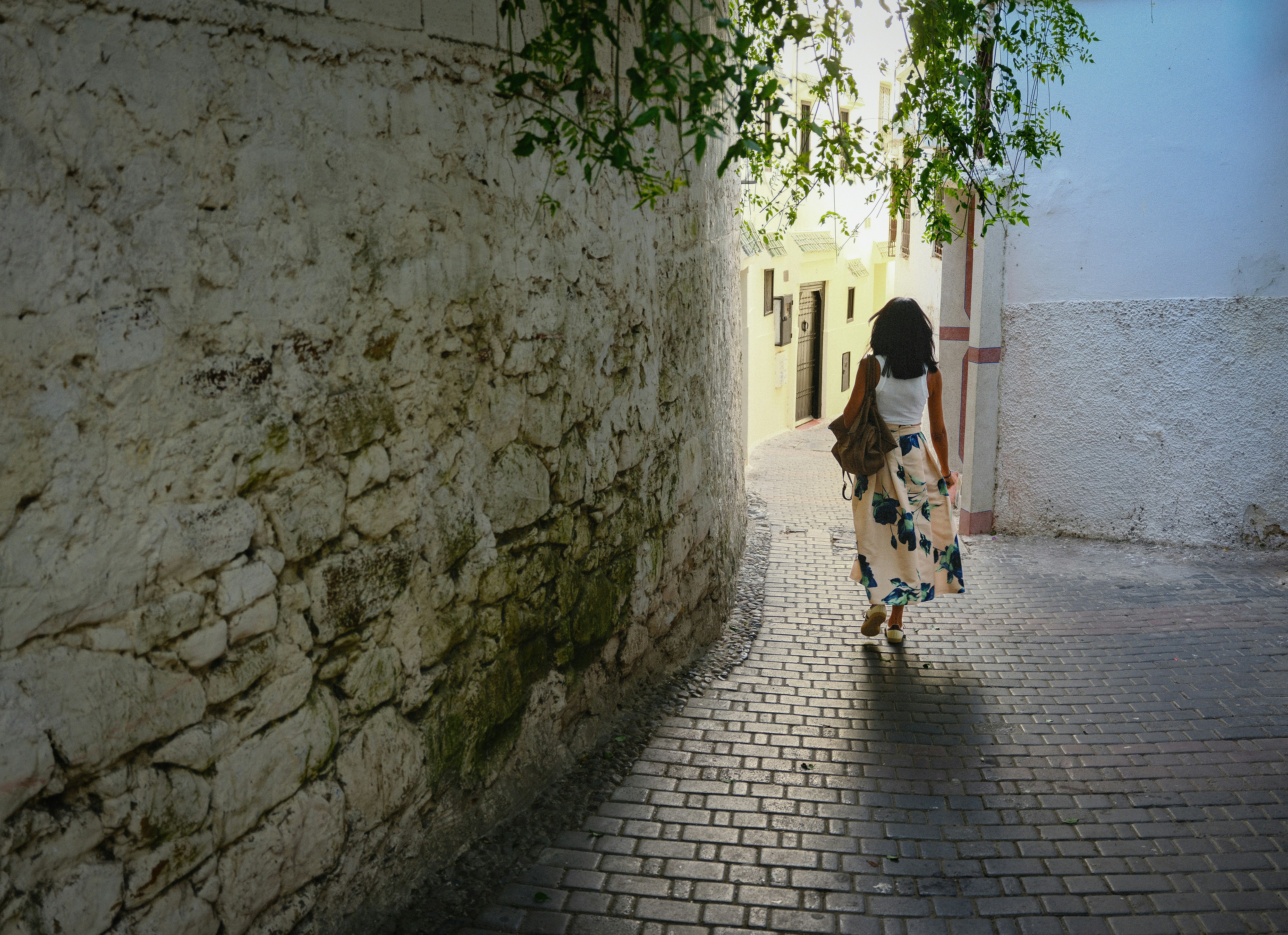 Ruelle paisible près de la medina de Tanger, au Maroc : pavés, murs blanchis et lumière douce, feuillage, une femme s’éloigne vers la rue.