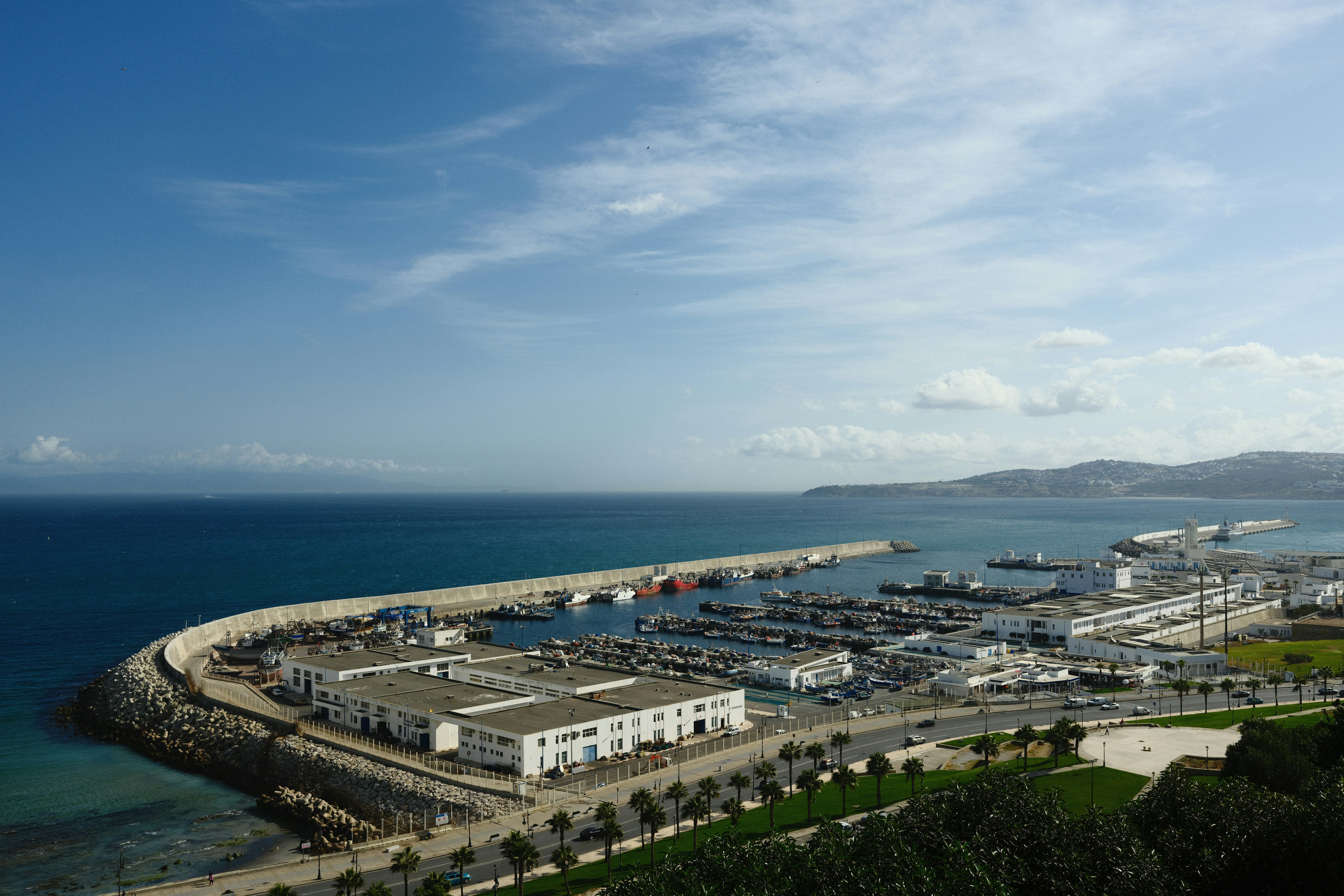 Vue panoramique du port de Tanger, au Maroc, avec sa digue courbe, ses bateaux de pêche et ses quais, bordés par une mer bleu profond sous un grand ciel. Une scène maritime d’Afrique du Nord.