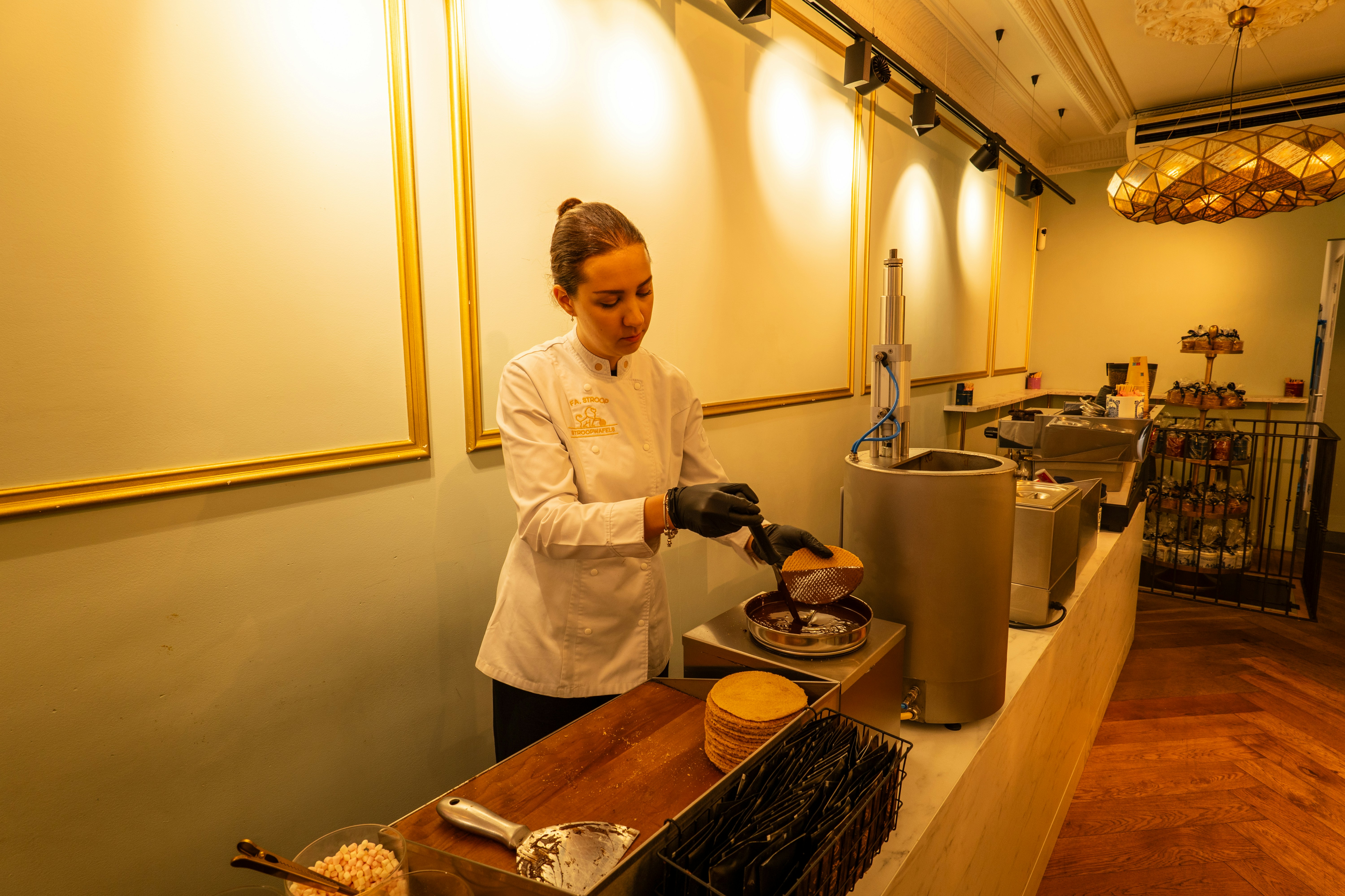 A warm and inviting scene inside a traditional Dutch stroopwafel shop in Amsterdam. A pastry chef carefully coats freshly baked waffle cookies with melted chocolate, while customers browse shelves filled with packaged stroopwafels and sweet treats. The elegant interior, golden lighting, and visible craftsmanship highlight the artisanal side of Dutch street food culture, captured in the heart of the city.