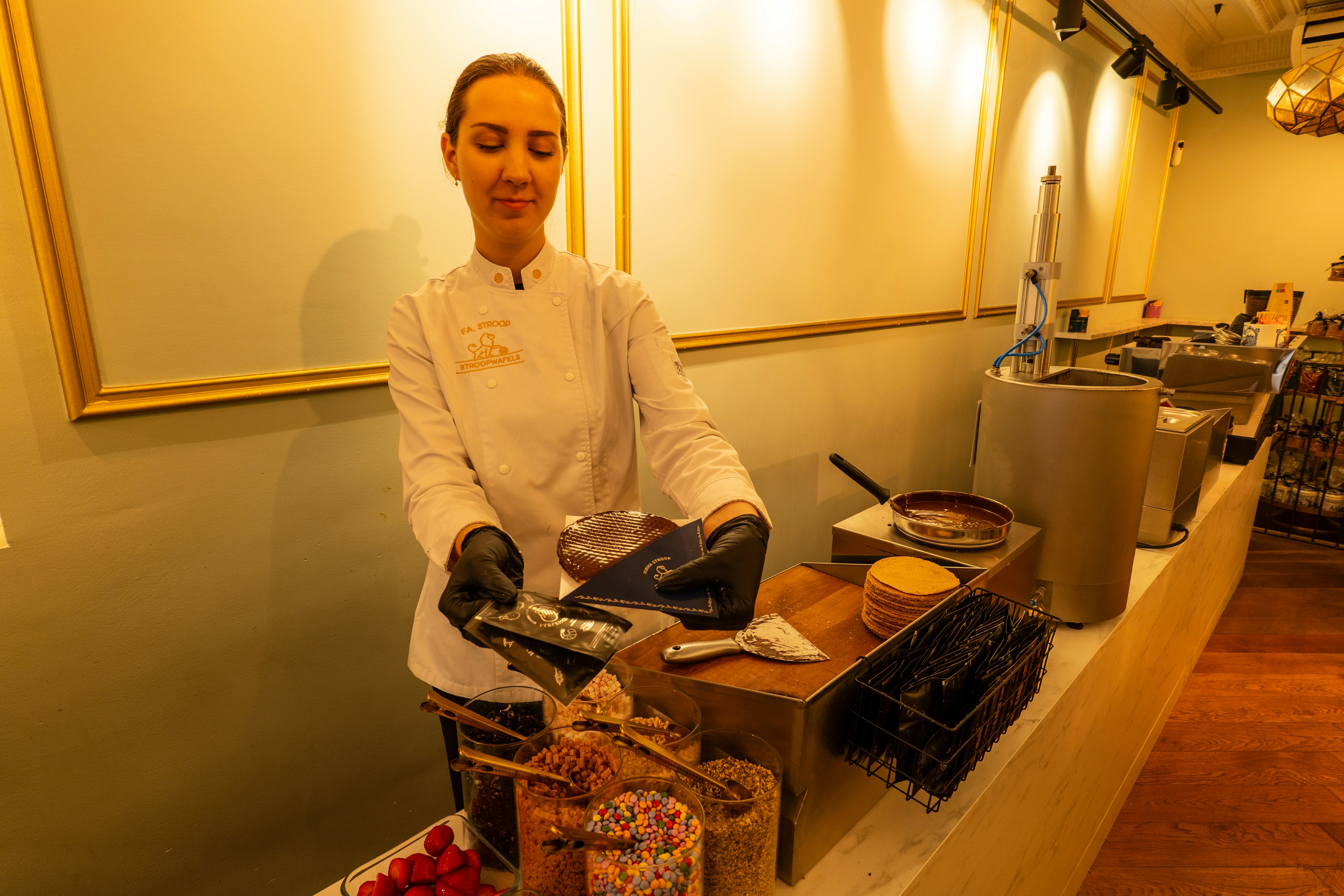 A warm and inviting scene inside a traditional Dutch stroopwafel shop in Amsterdam. A pastry chef carefully coats freshly baked waffle cookies with melted chocolate, while customers browse shelves filled with packaged stroopwafels and sweet treats. The elegant interior, golden lighting, and visible craftsmanship highlight the artisanal side of Dutch street food culture, captured in the heart of the city.