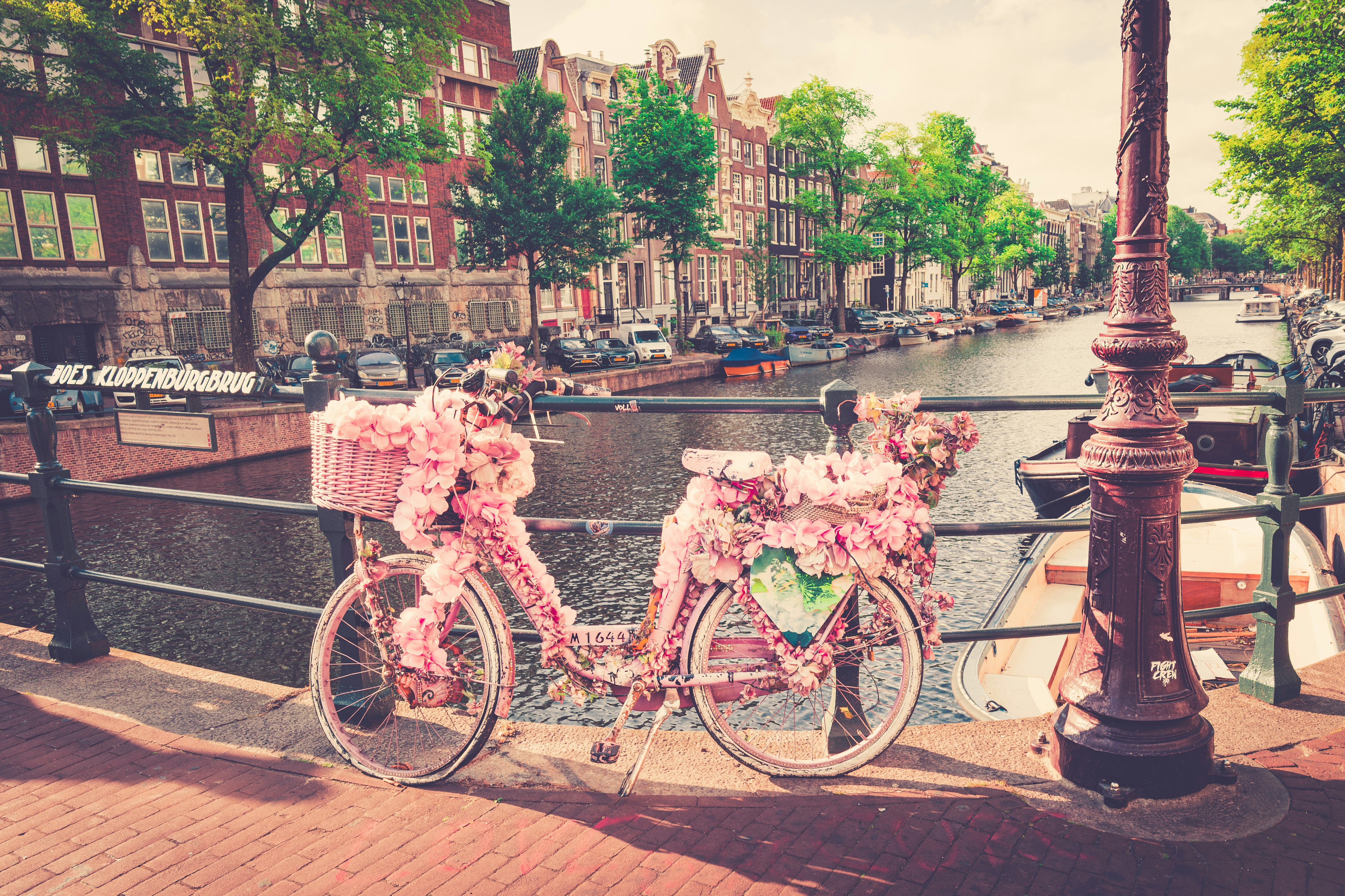 Decorated bicycles covered in flowers locked along a canal railing at Joos Kloppenburgbrug on the Keizersgracht in Amsterdam. The scene captures the city’s iconic blend of cycling culture, historic canal houses, and everyday urban poetry. Soft pastel tones, reflections on the water, and pedestrians crossing the bridge create a calm, romantic atmosphere typical of Amsterdam’s historic center.