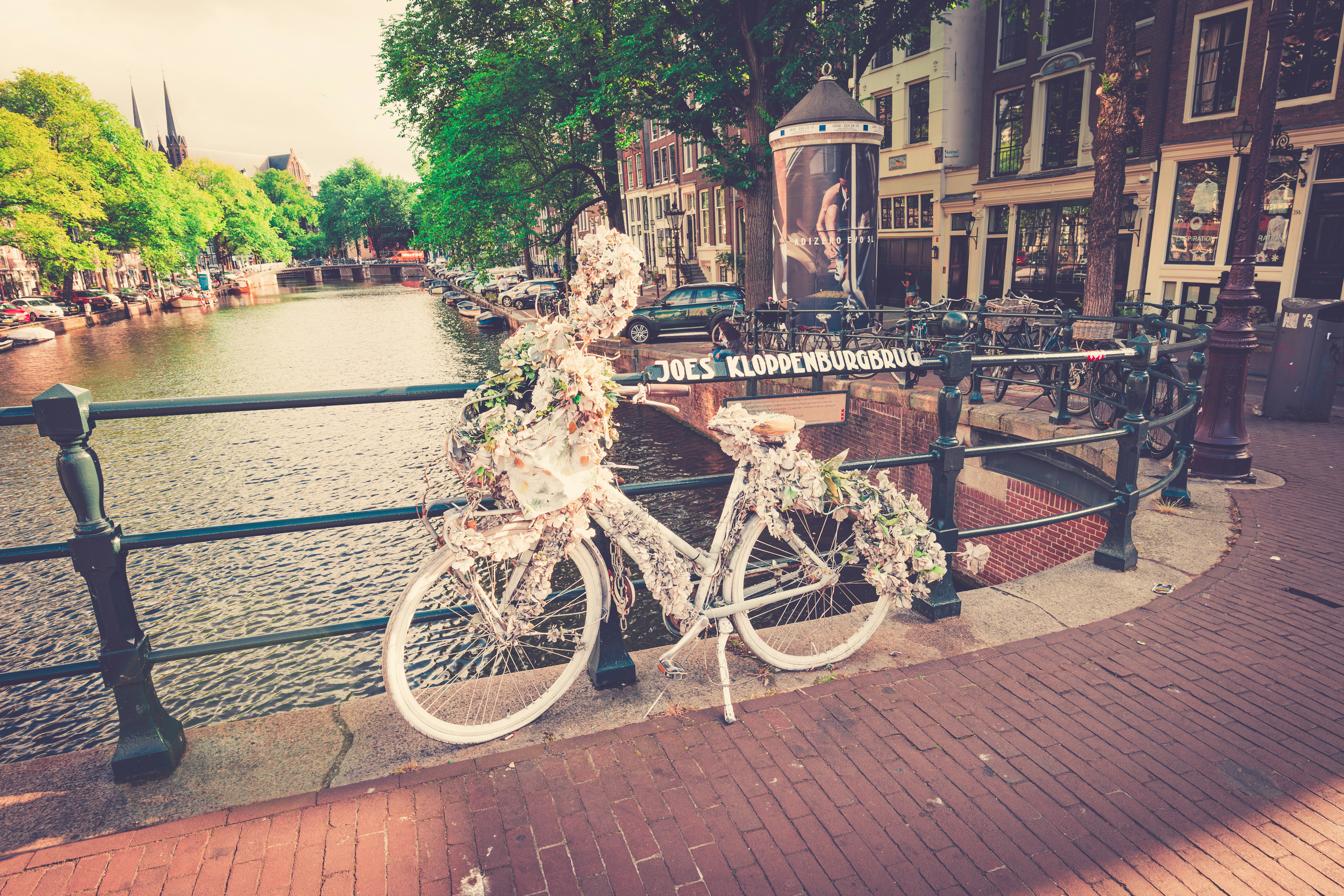 Decorated bicycles covered in flowers locked along a canal railing at Joos Kloppenburgbrug on the Keizersgracht in Amsterdam. The scene captures the city’s iconic blend of cycling culture, historic canal houses, and everyday urban poetry. Soft pastel tones, reflections on the water, and pedestrians crossing the bridge create a calm, romantic atmosphere typical of Amsterdam’s historic center.