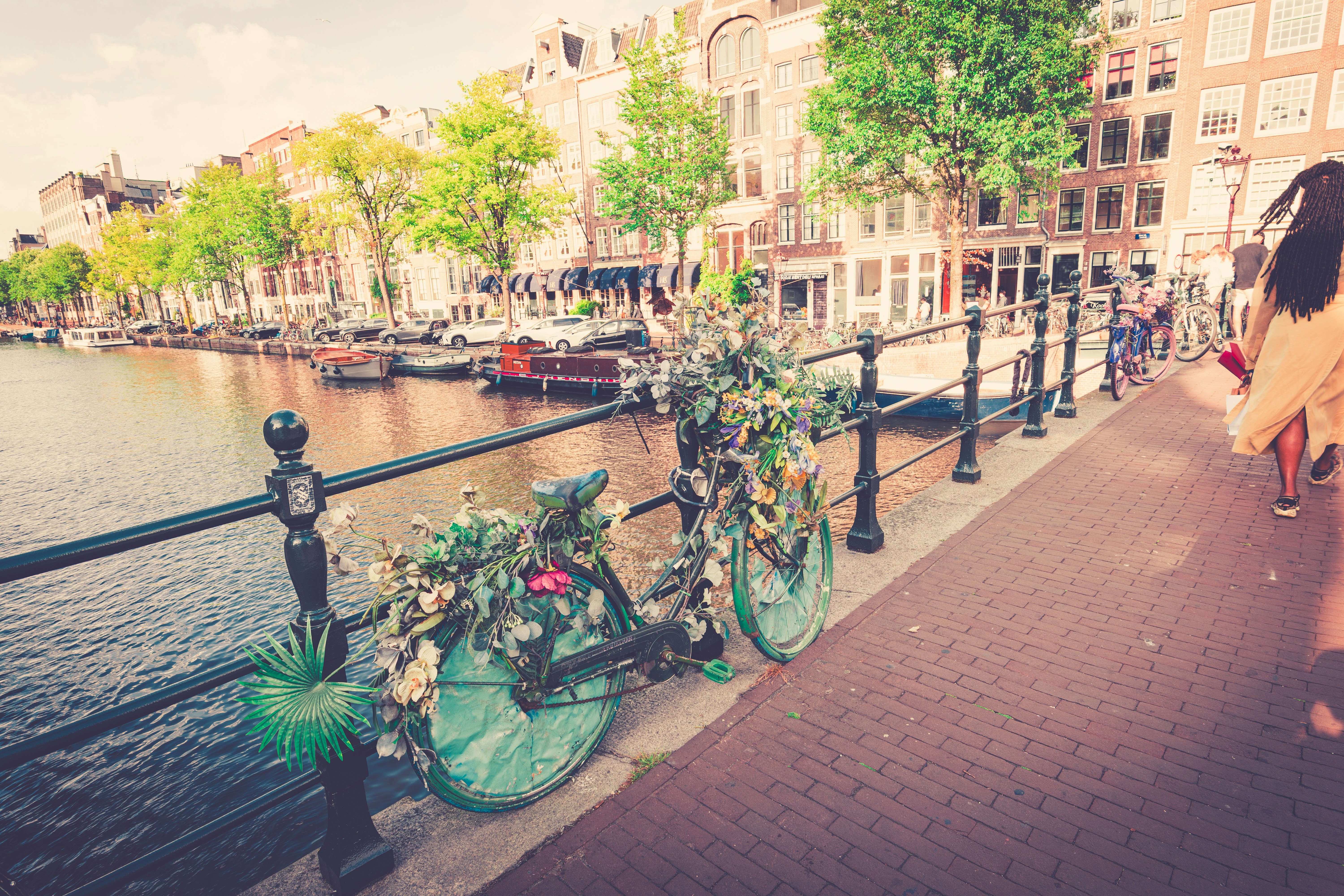 Decorated bicycles covered in flowers locked along a canal railing at Joos Kloppenburgbrug on the Keizersgracht in Amsterdam. The scene captures the city’s iconic blend of cycling culture, historic canal houses, and everyday urban poetry. Soft pastel tones, reflections on the water, and pedestrians crossing the bridge create a calm, romantic atmosphere typical of Amsterdam’s historic center.