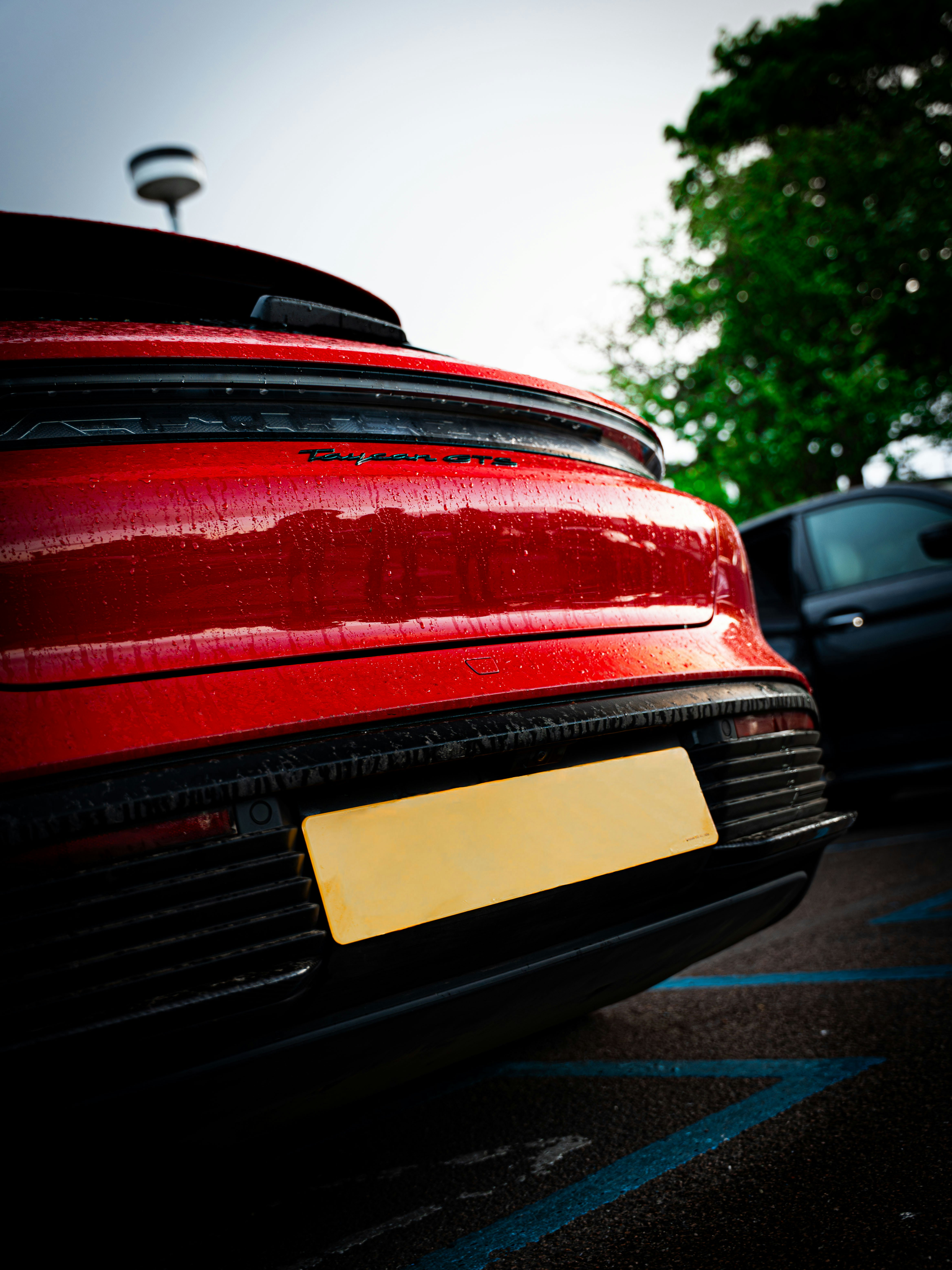 Close-up of the rear end of a red car with weathered paint and a yellow license plate in a parking lot, showcasing glossy curves and reflections.