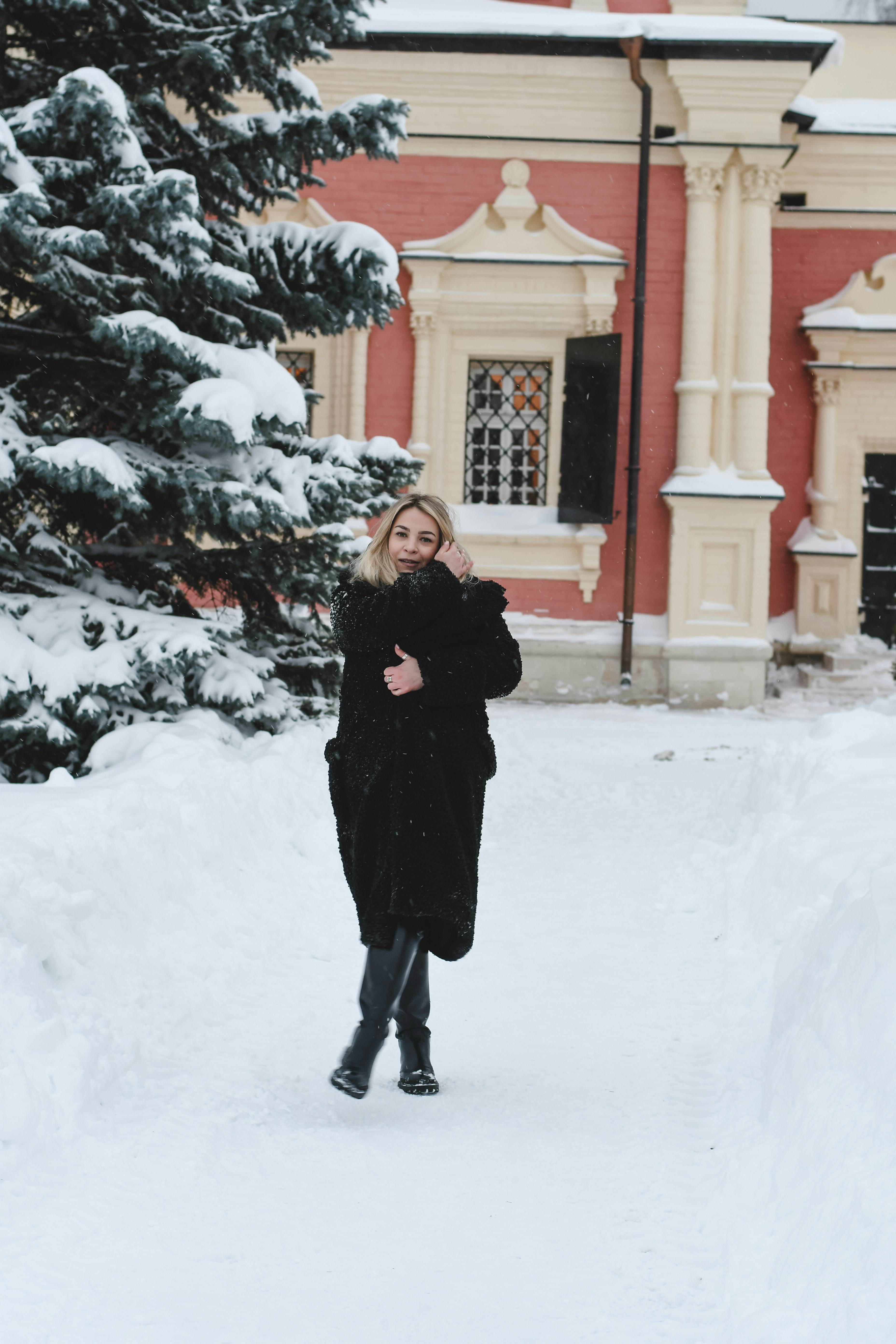 A woman in a stylish long black coat walking through a snow-covered path. The scene features a beautiful contrast between the dark winter attire, the white snow, and a classic red building with ornate white details in the background. A snow-laden evergreen tree adds to the serene, wintry atmosphere.