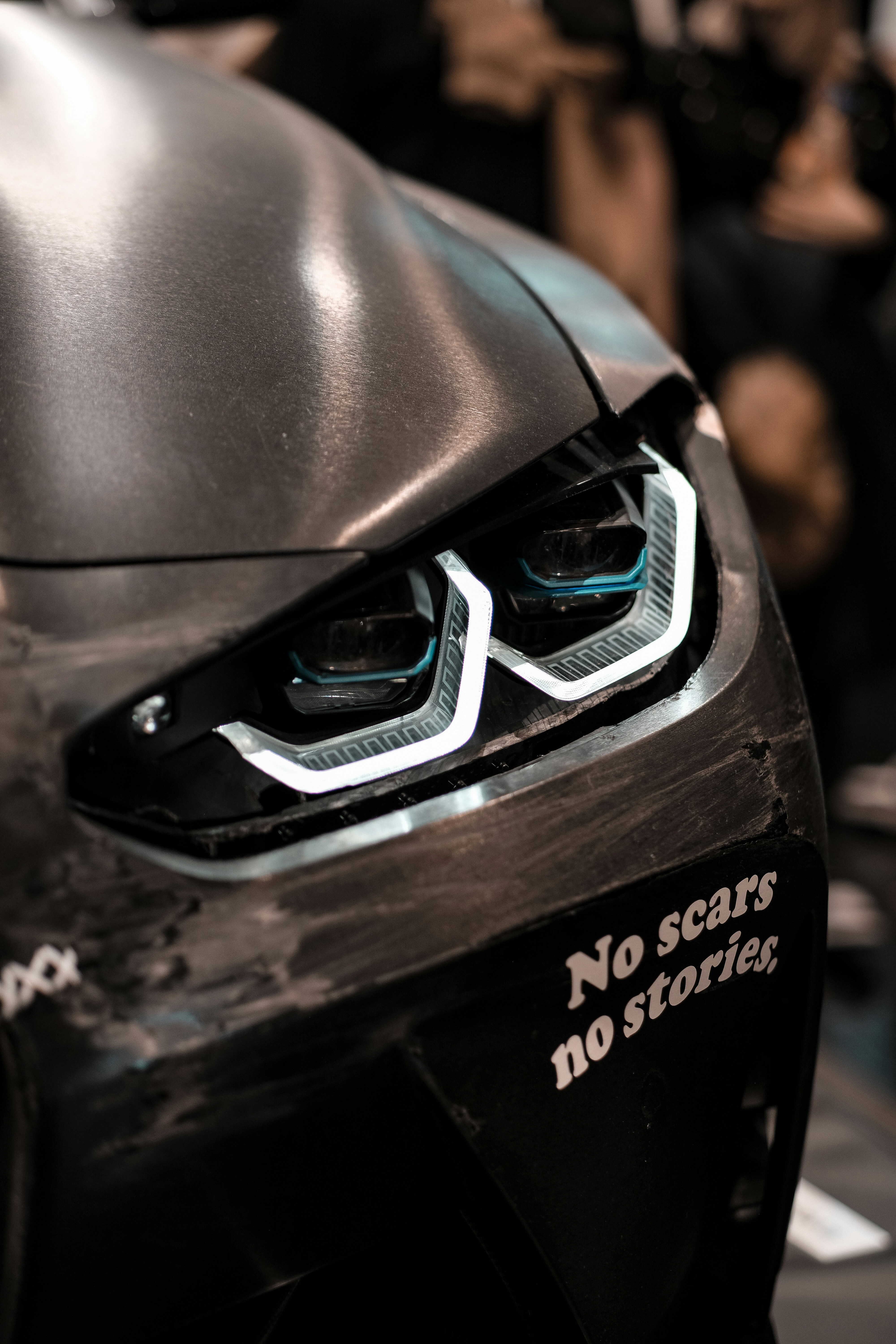 Close-up of a sports car’s aggressive headlight with blue-tinted elements; the surrounding metallic body shows a racing-inspired finish and a partial logo on the bumper.
