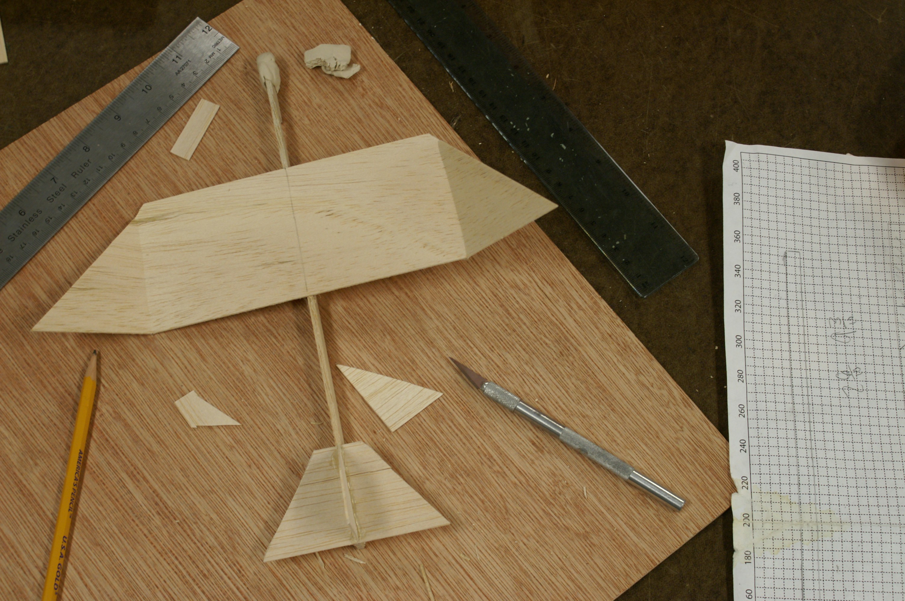A balsa wood glider on a workbench with tools and graph paper scattered around it.