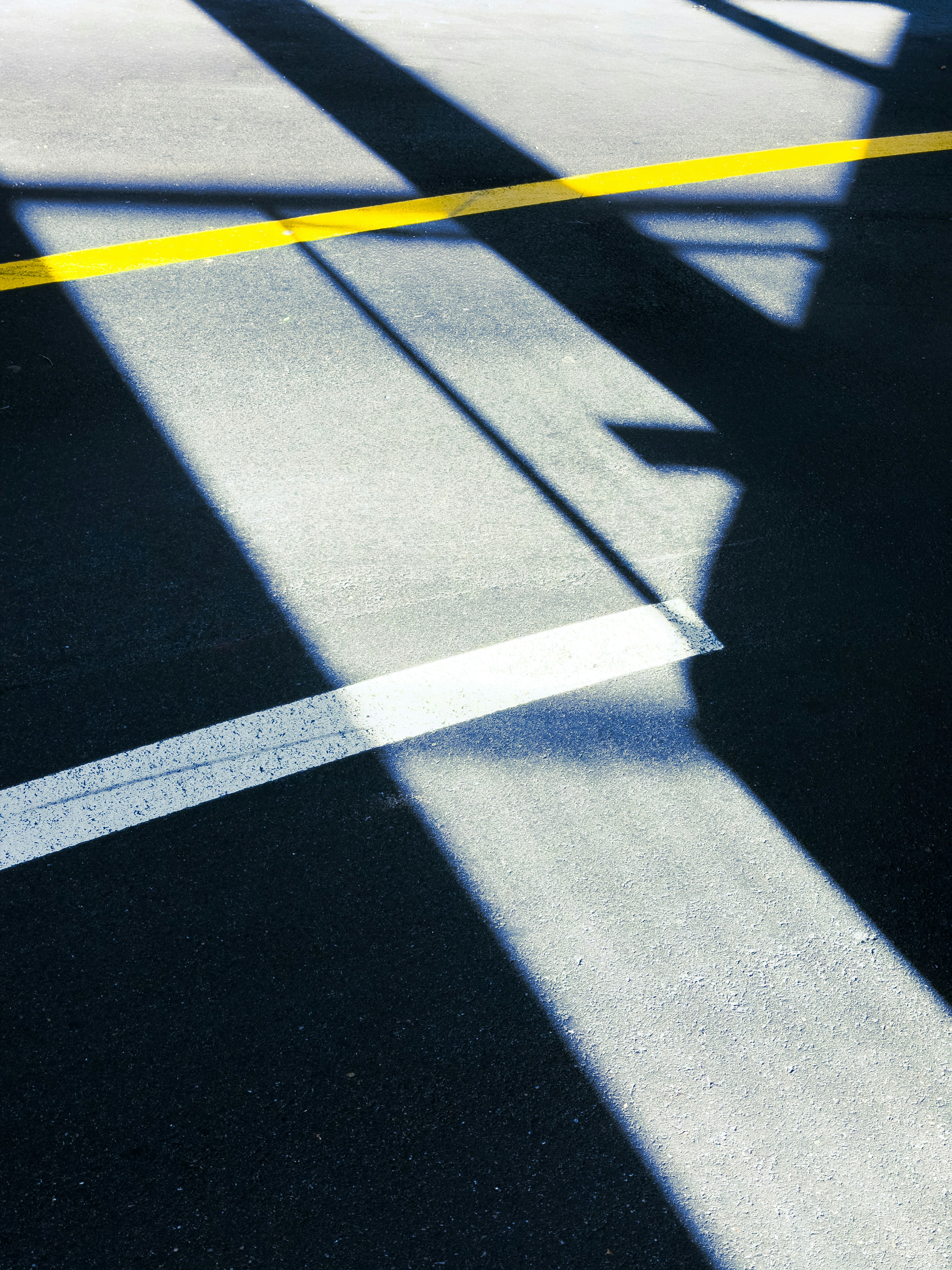 Shadow shapes and colored lines cast across a dark asphalt parking area, creating angular patterns and a sense of depth. A bright yellow line slices through the composition, emphasizing geometry and contrast.
