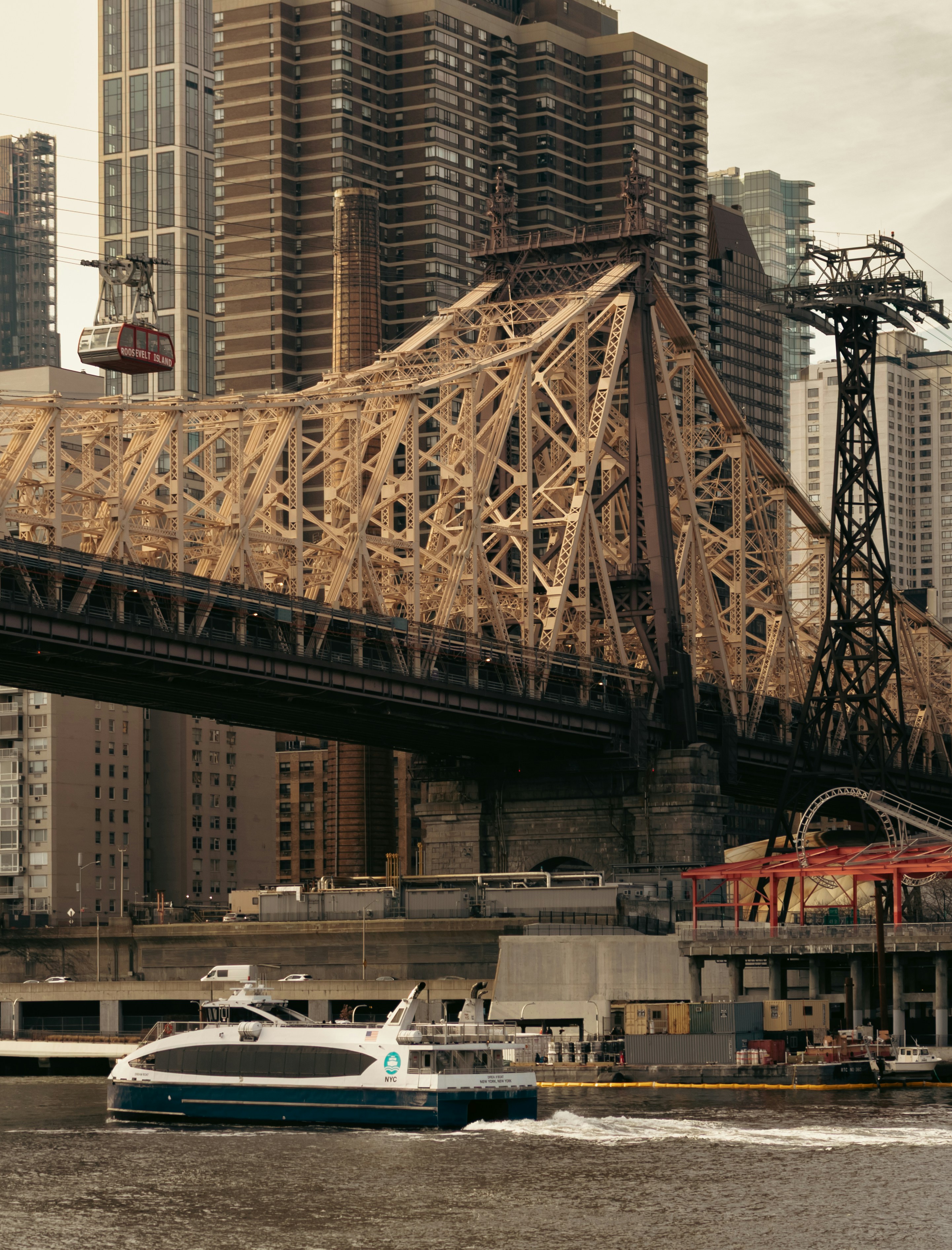 Riverfront scene with a large steel truss bridge spanning a calm waterway, framed by high-rise buildings; a white-and-blue boat cruises in the foreground, creating a contrast between architecture and movement.