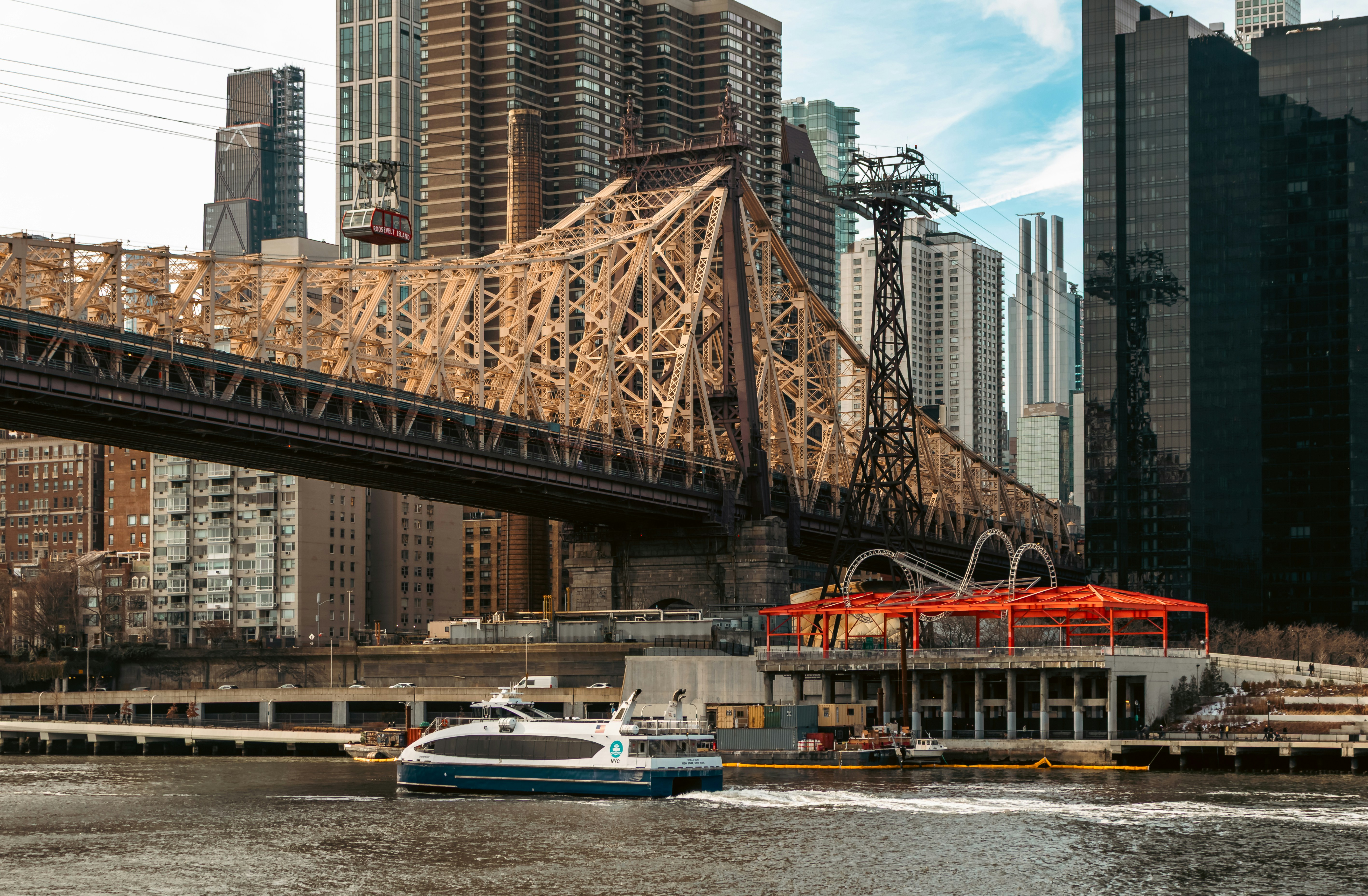 A riverfront scene featuring a lattice steel bridge with towering skyscrapers in the background and a white-and-blue boat docked near a red-roofed terminal at the water's edge.