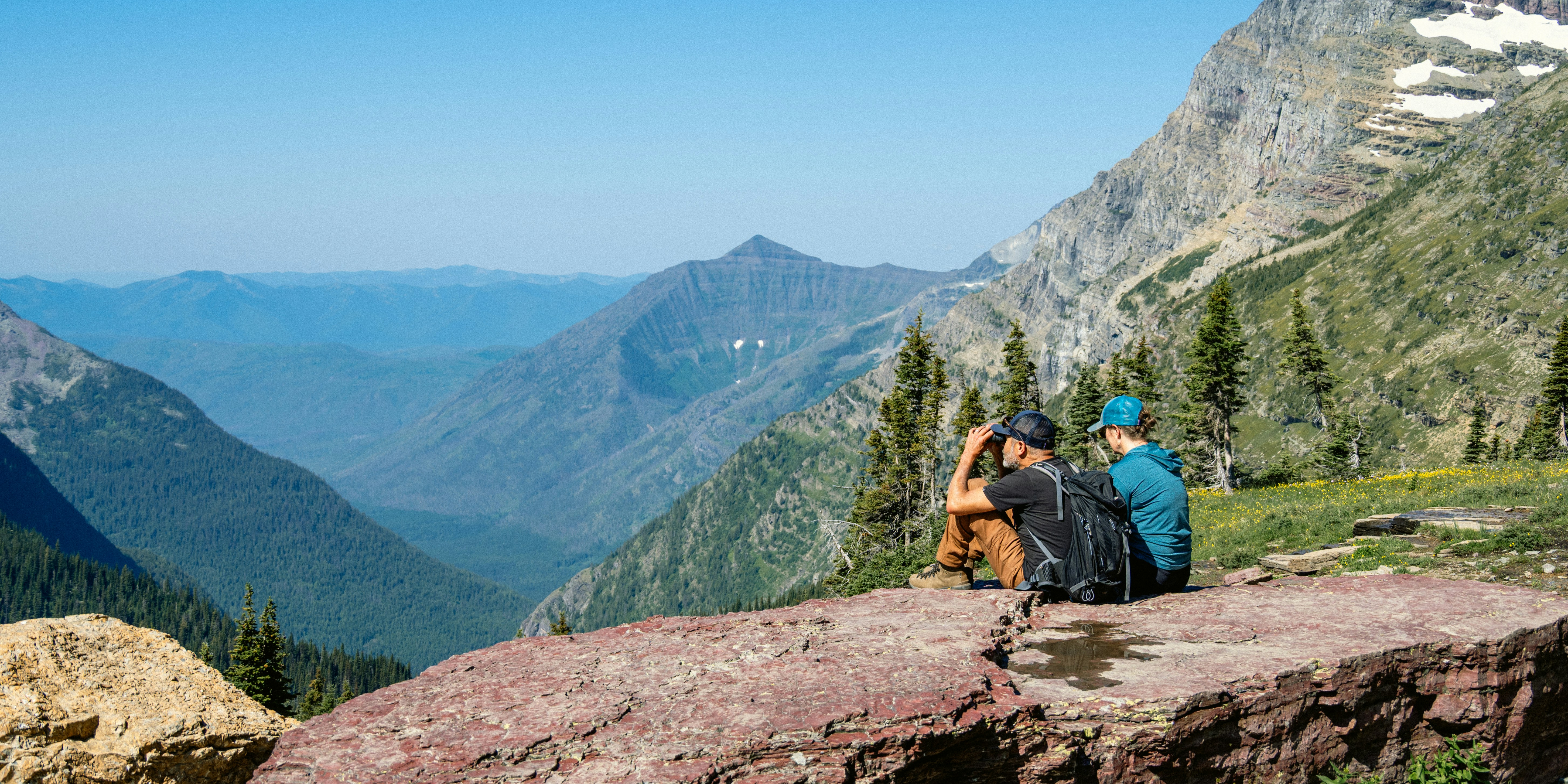Two hikers on a sunlit cliff overlook with distant mountain silhouettes