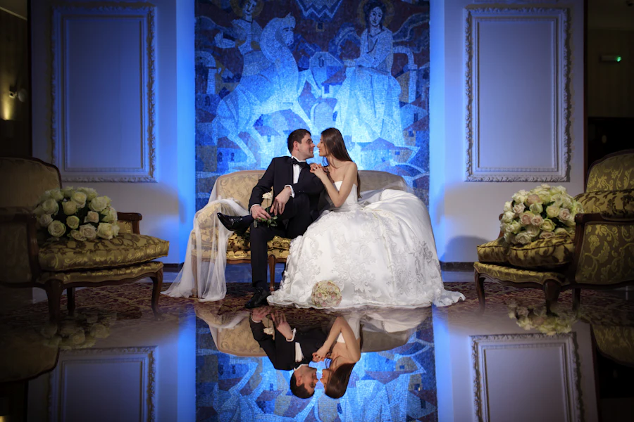 Bride and groom share a kiss on a lavish thronelike couch, blue ambient lighting highlights a grand mural, their reflection glimmering in the mirrored floor.