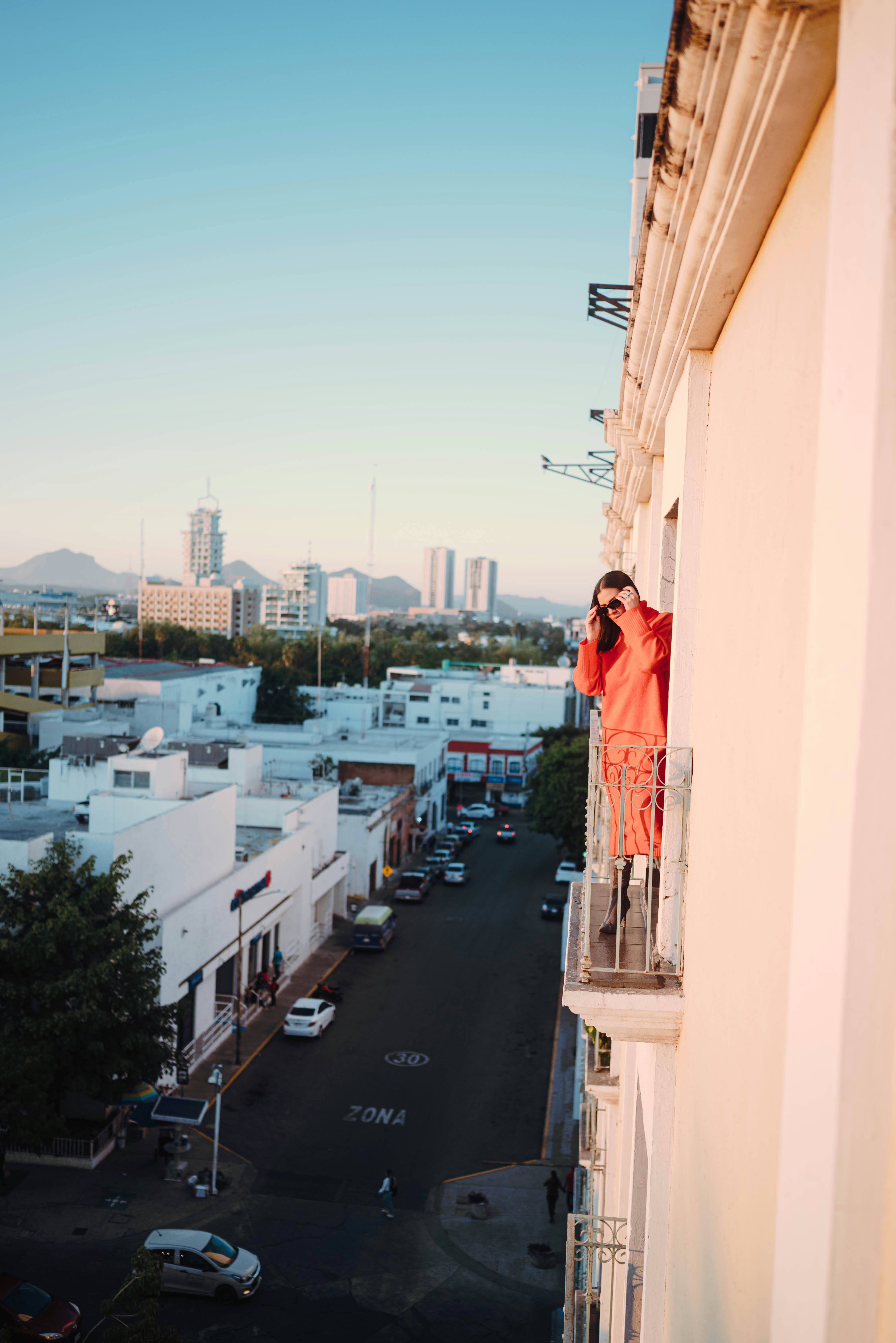 Edge of the Balcony: A Photographer on a Coral Orange Outfit Peering Over the City