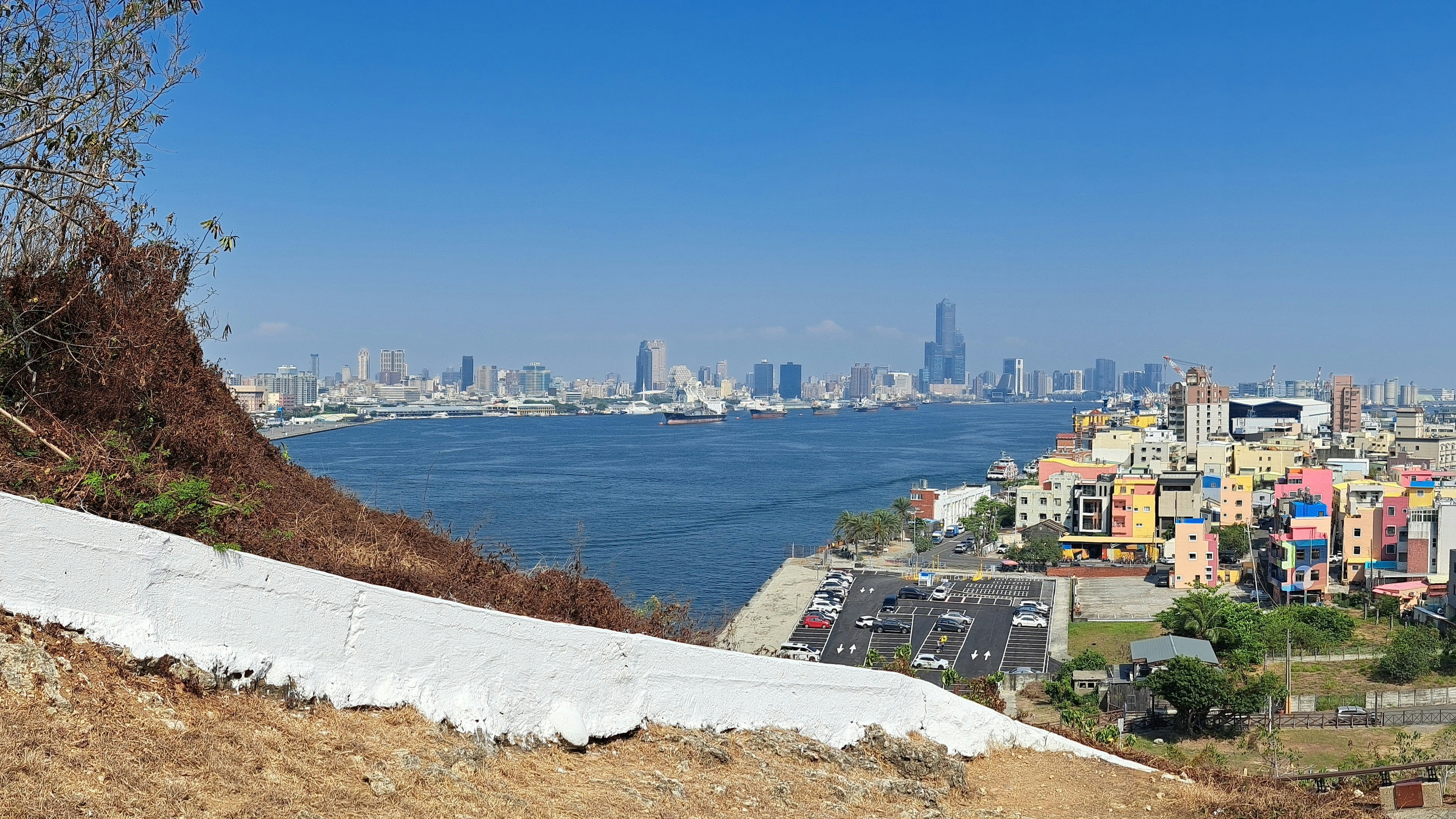 Coastal park overlooks a vibrant cityscape across a calm bay, with a mix of colorful buildings lining the waterfront.