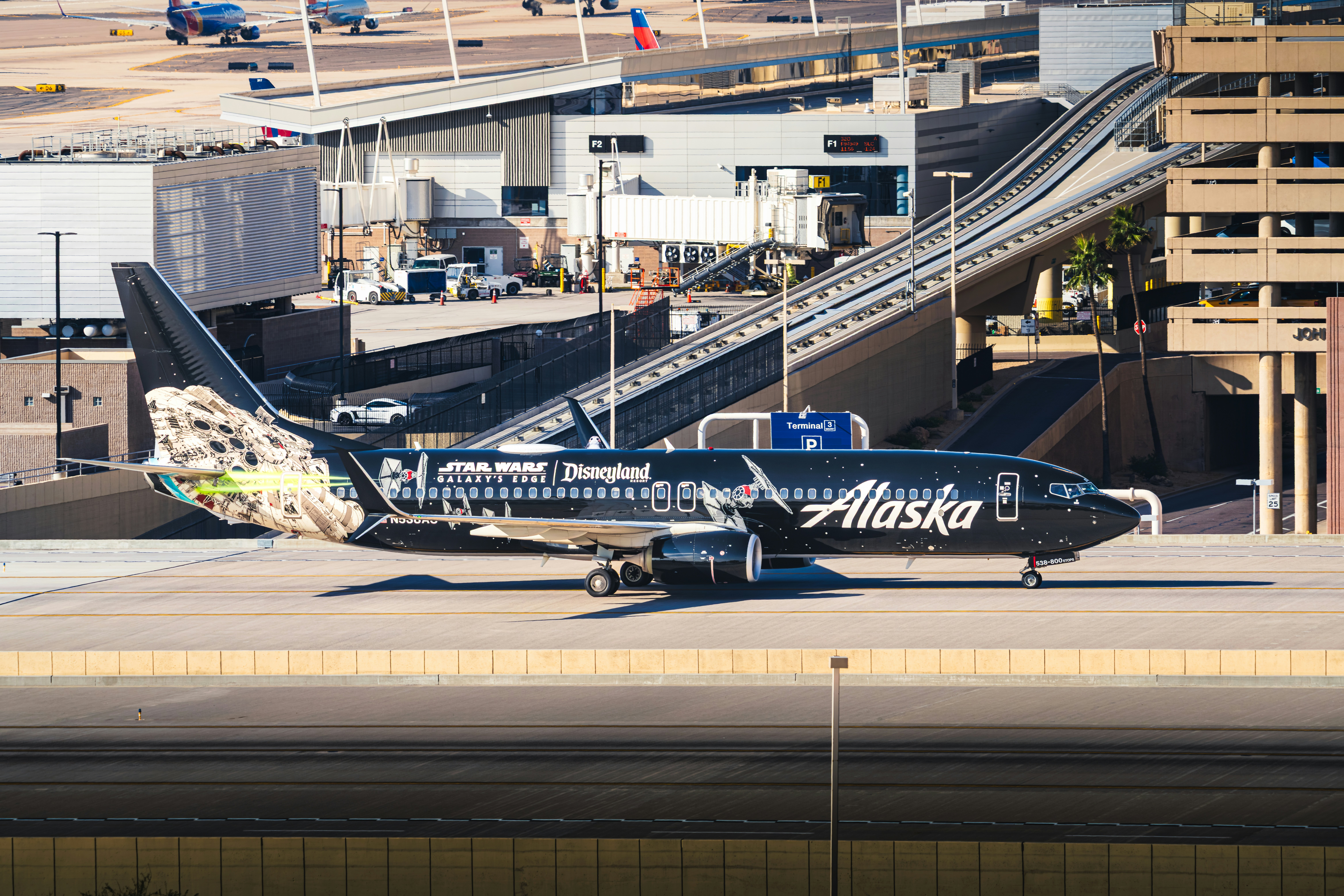 Alaska Airlines A320neo taxiing past elevated walkway at a sunlit terminal