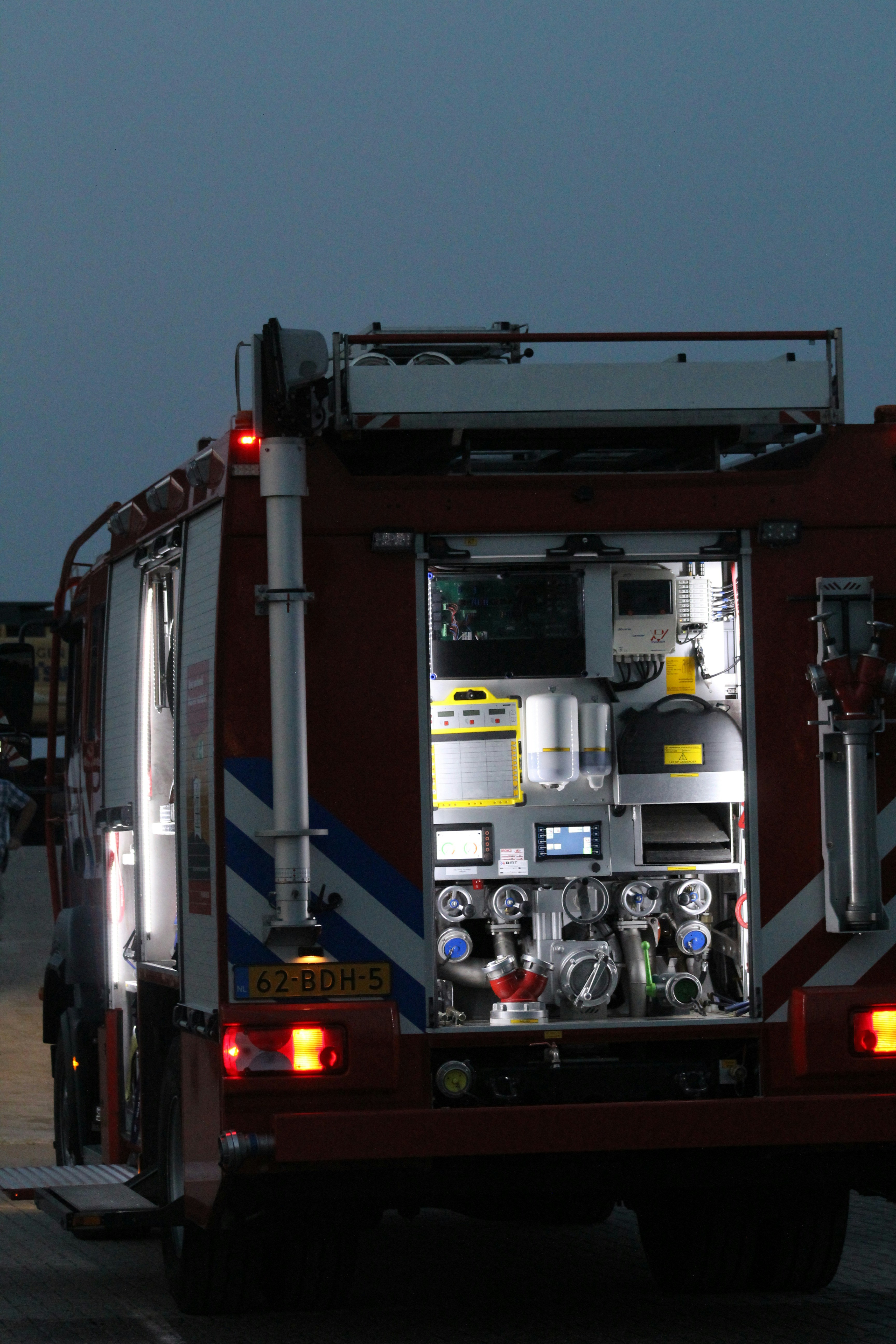 Fire engine with open rear cabinet showing hoses and gauges; subtle dusk lighting highlights the organized interior and red exterior.