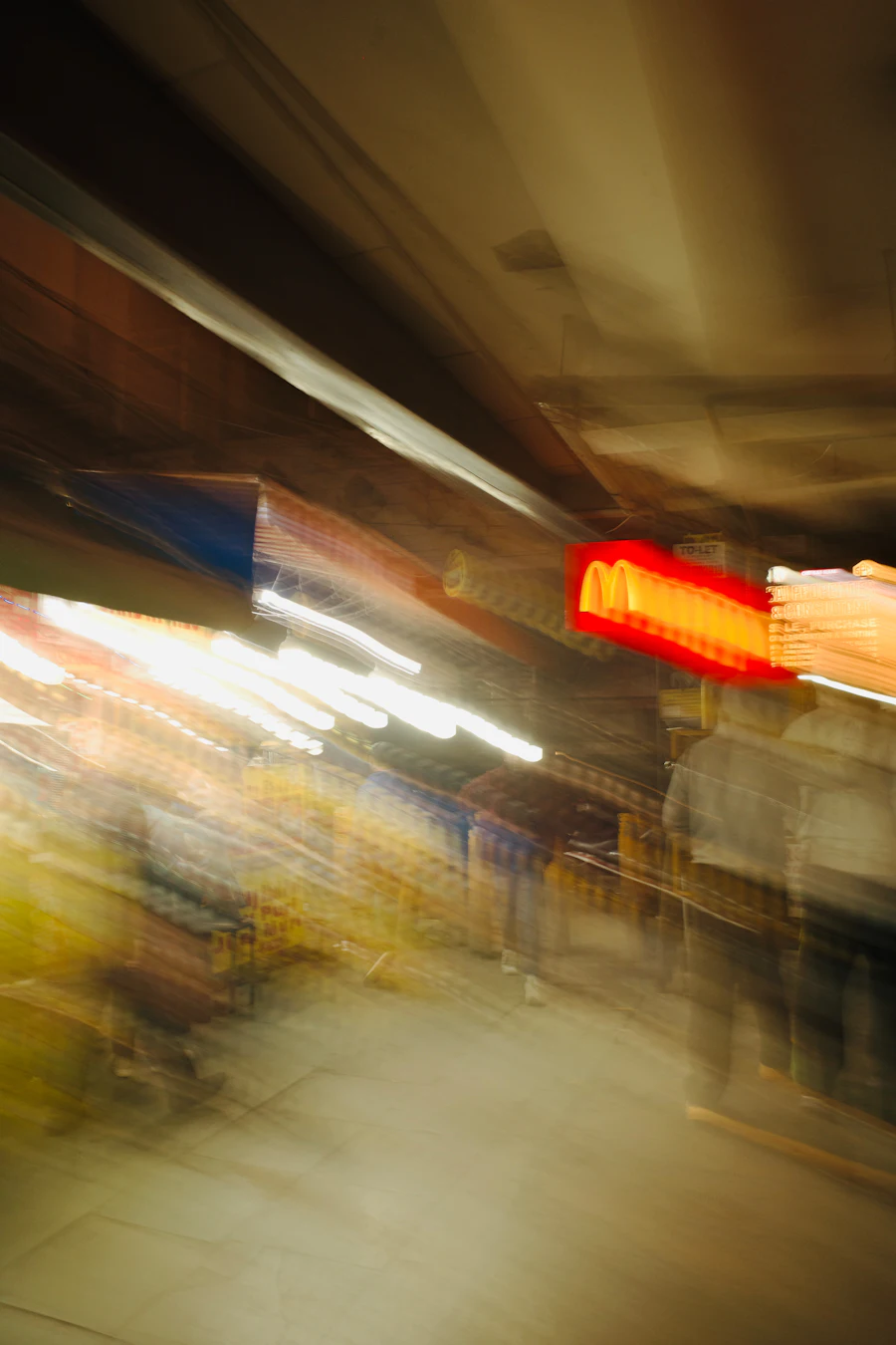 Motion-blurred urban street scene with a glowing McDonald's sign as a bright focal point amid streaks of lights and hazy figures.