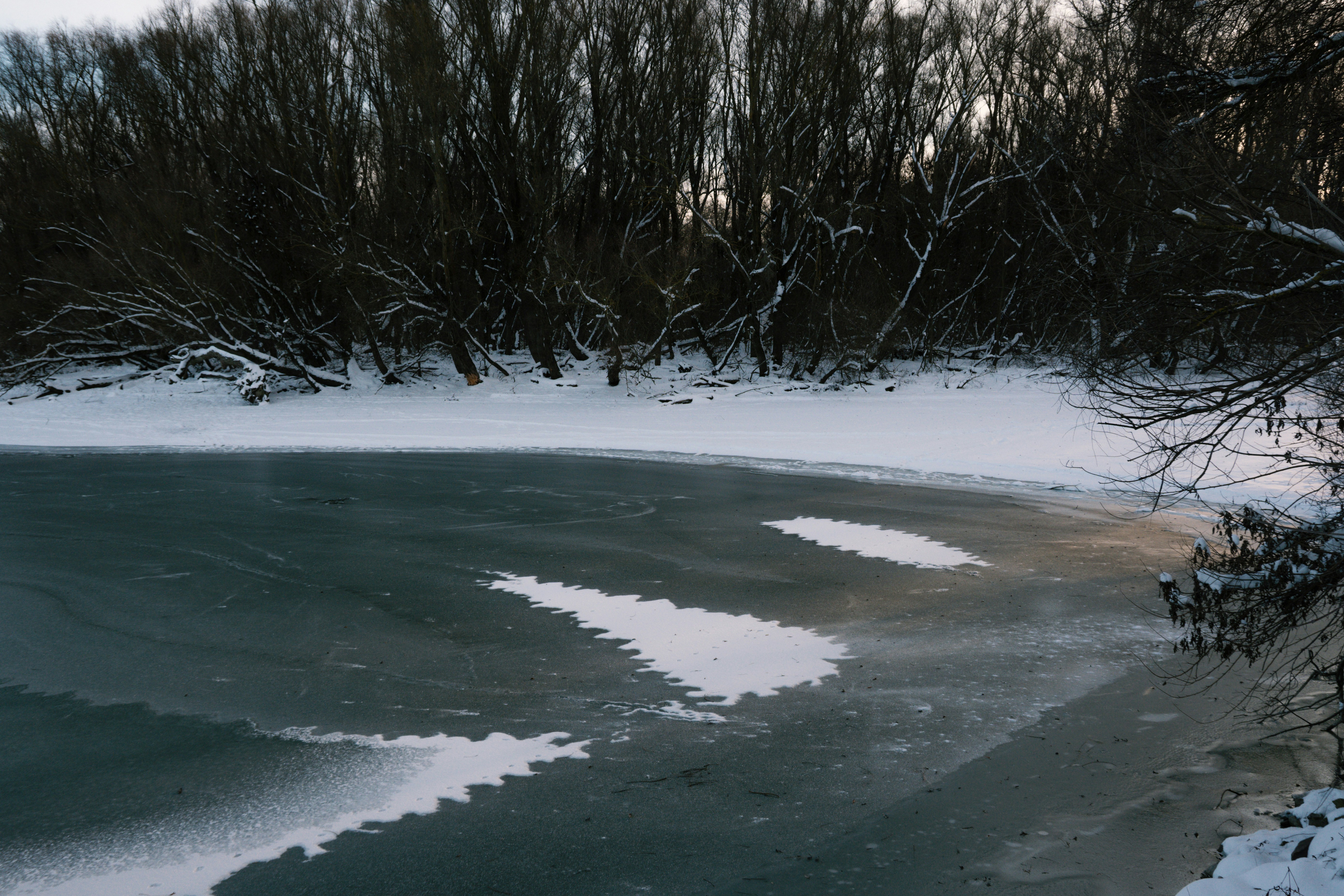 Icy shoreline with dark snow-dusted trees framing a calm, partially frozen waterway and irregular white ice patches across the surface. A wintry landscape scene with muted colors and a quiet mood.