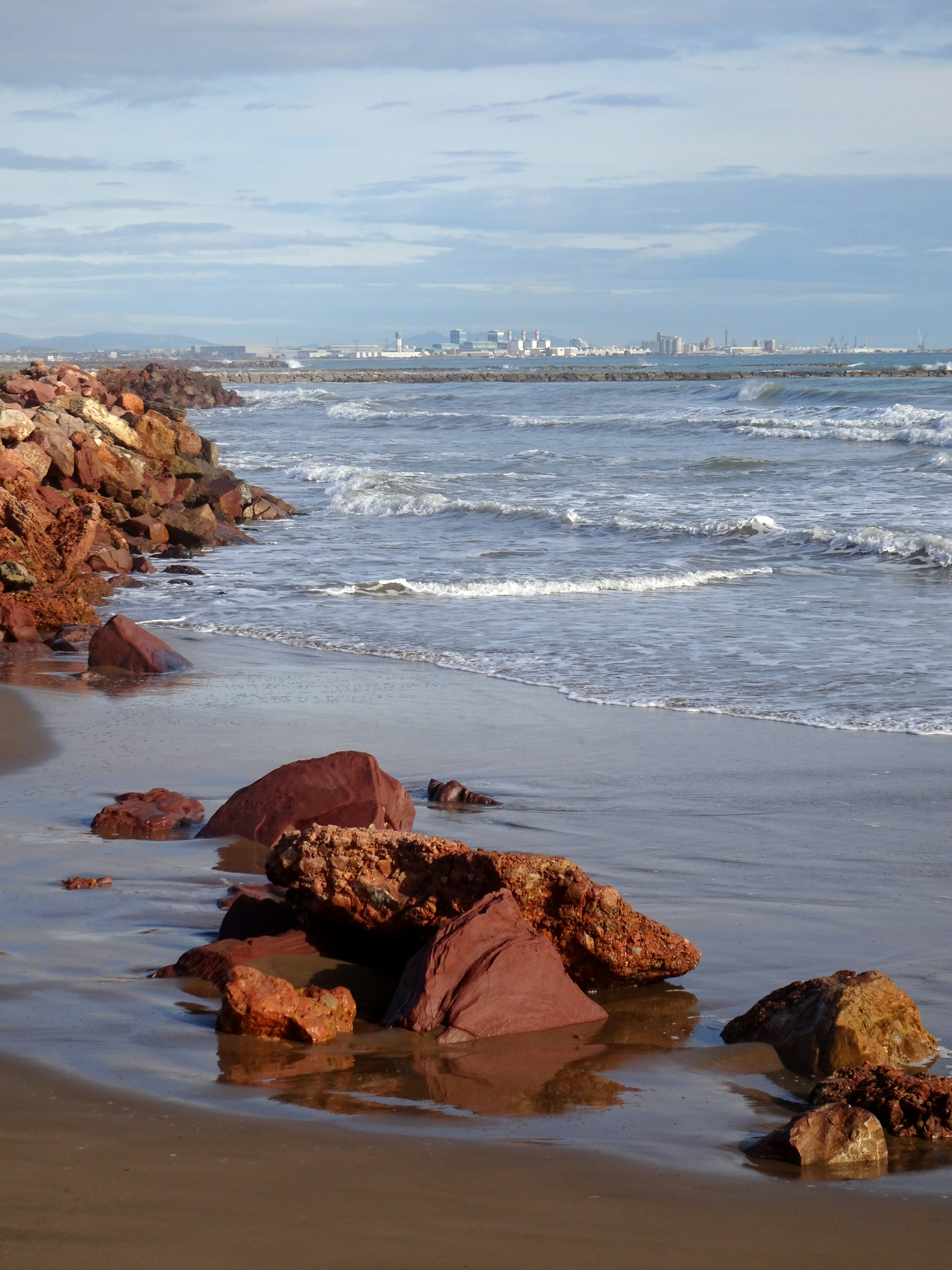 Paisaje de playa con unas rocas en primer plano y contrucciones al fondo.
