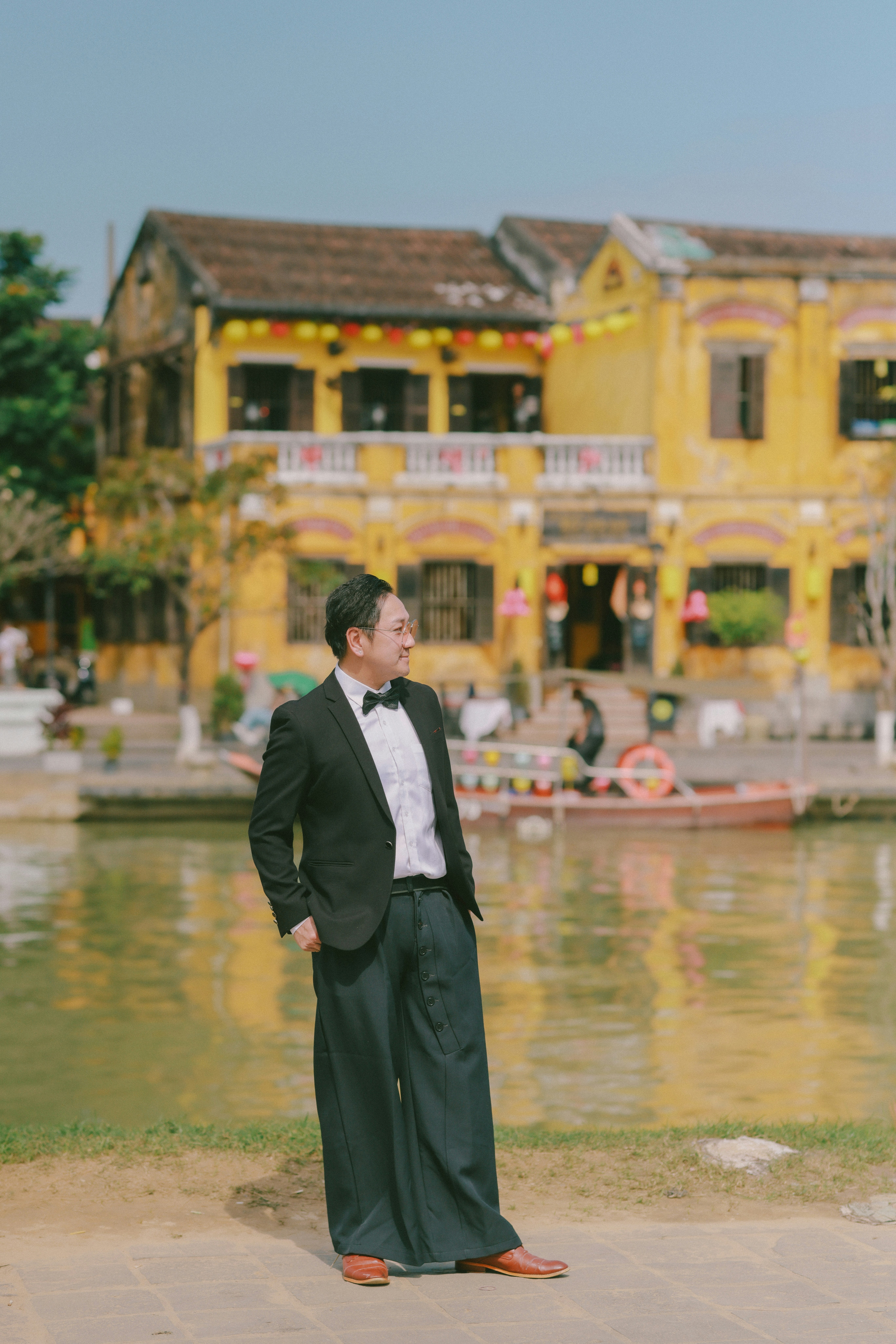 Groom in a tuxedo shirt stands by a sunlit riverside terrace