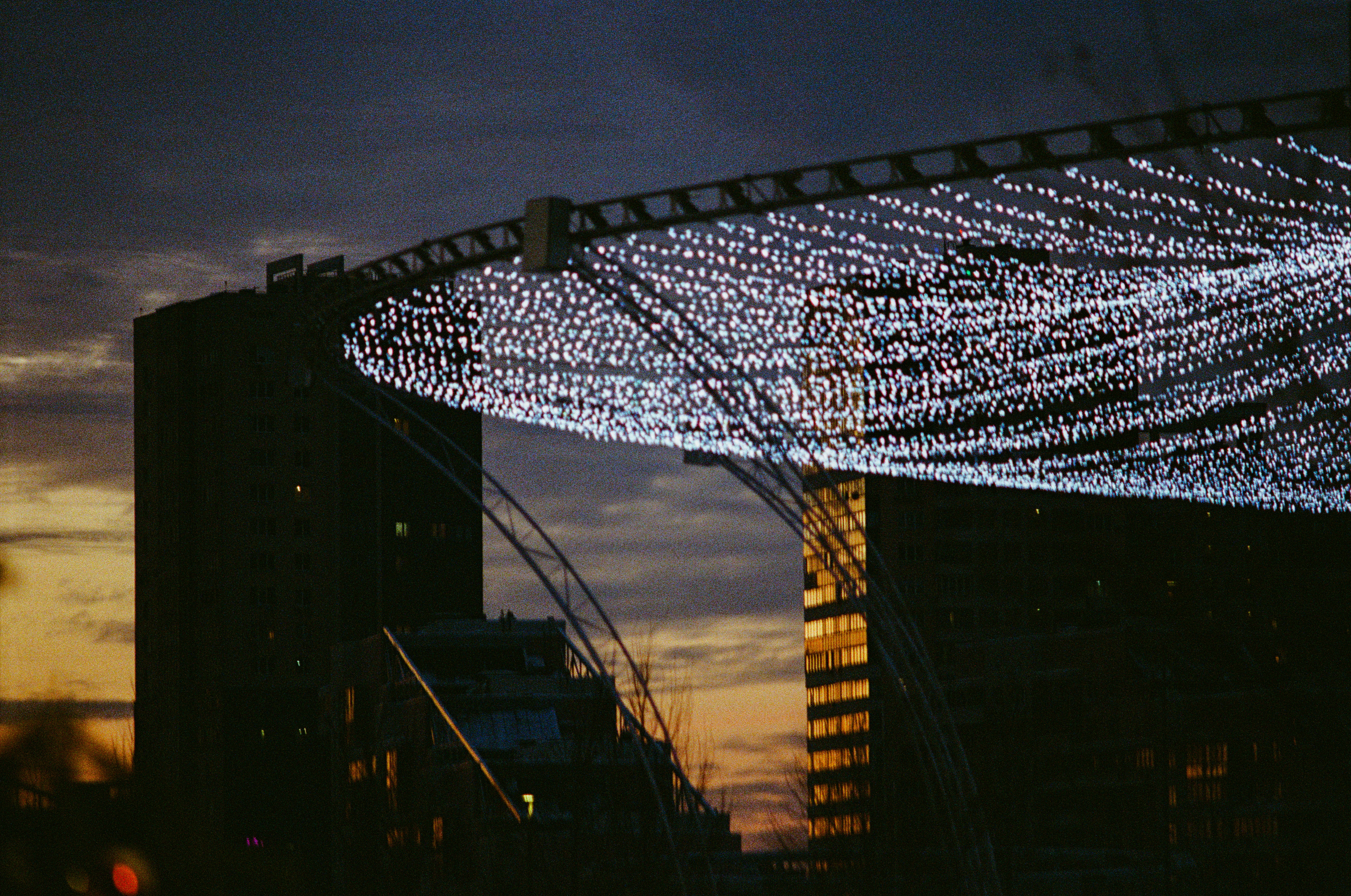 Urban evening cityscape featuring a decorative canopy of string lights against a sunset sky. Silhouettes of modern buildings in low light with film grain texture. Film: ISO 400 (+2 stops push) | Lens: Takumar 200mm f/4.0