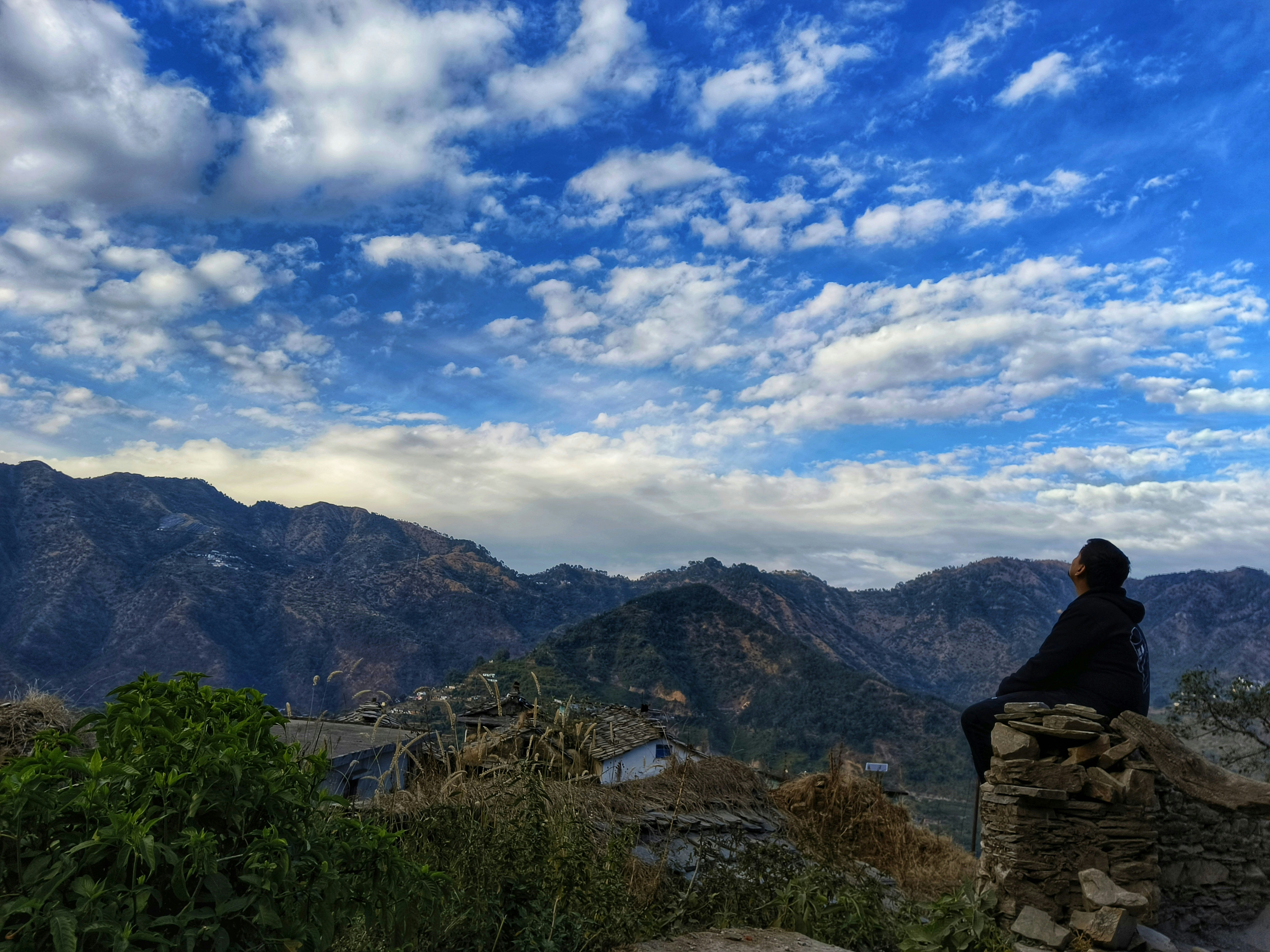 A person sitting on a stone wall overlooking a quiet mountain village, gazing at a vast blue sky filled with scattered clouds. The wide landscape and still moment convey solitude, reflection, and a deep connection with nature and travel.