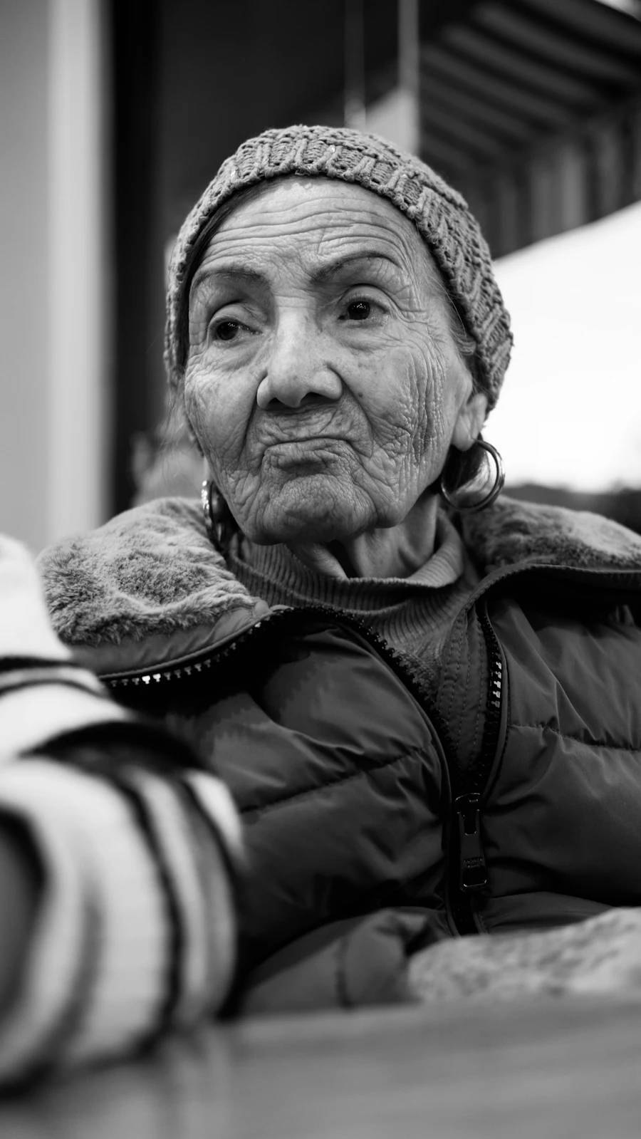 An elderly woman in a knit hat sits against a neutral background, her face lined with years and a subtle, contemplative expression. A rugged jacket and hoop earring add character; this is a candid, intimate portrait.