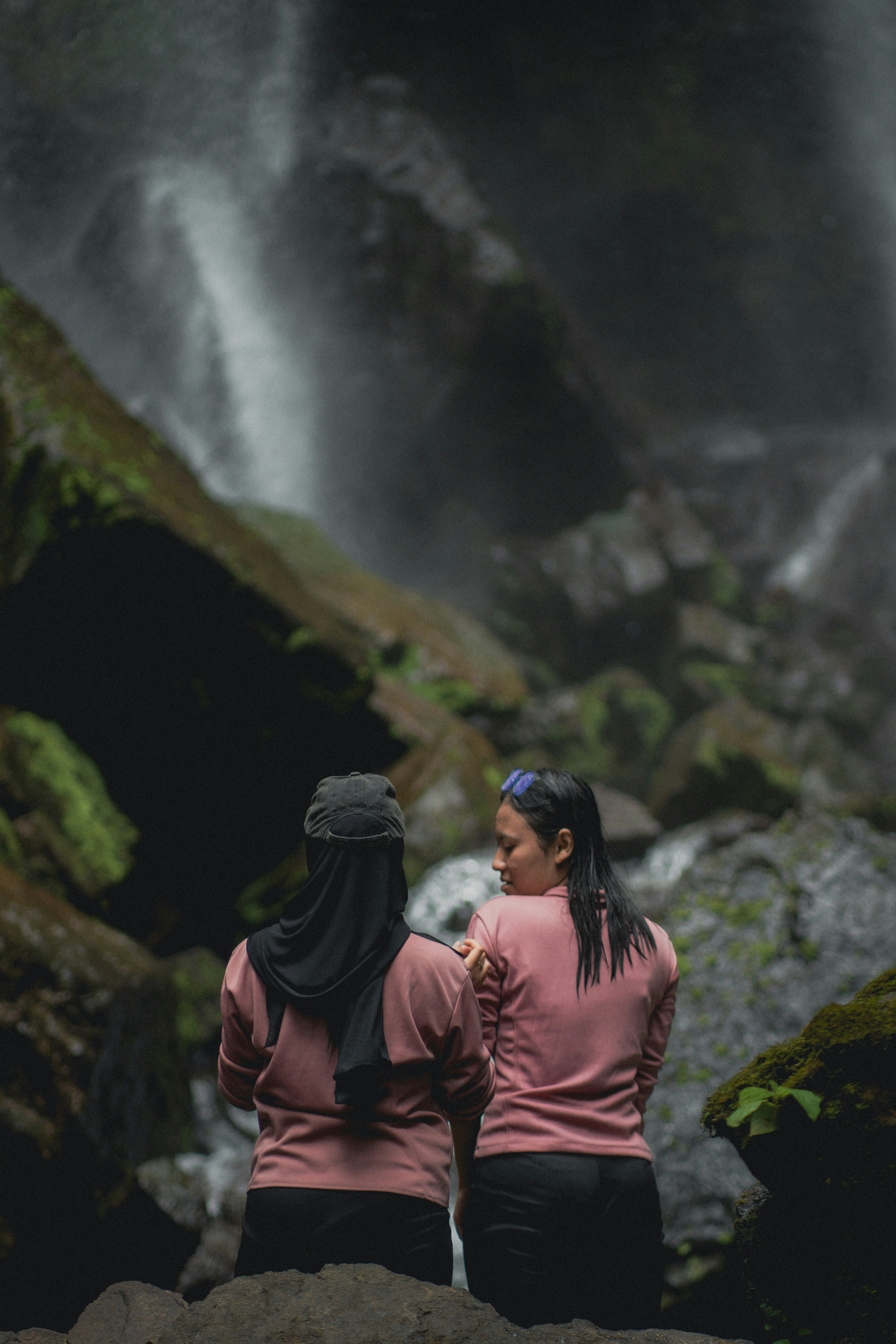 Two friends in soft pink jackets stand before a mossy cavern and waterfall
