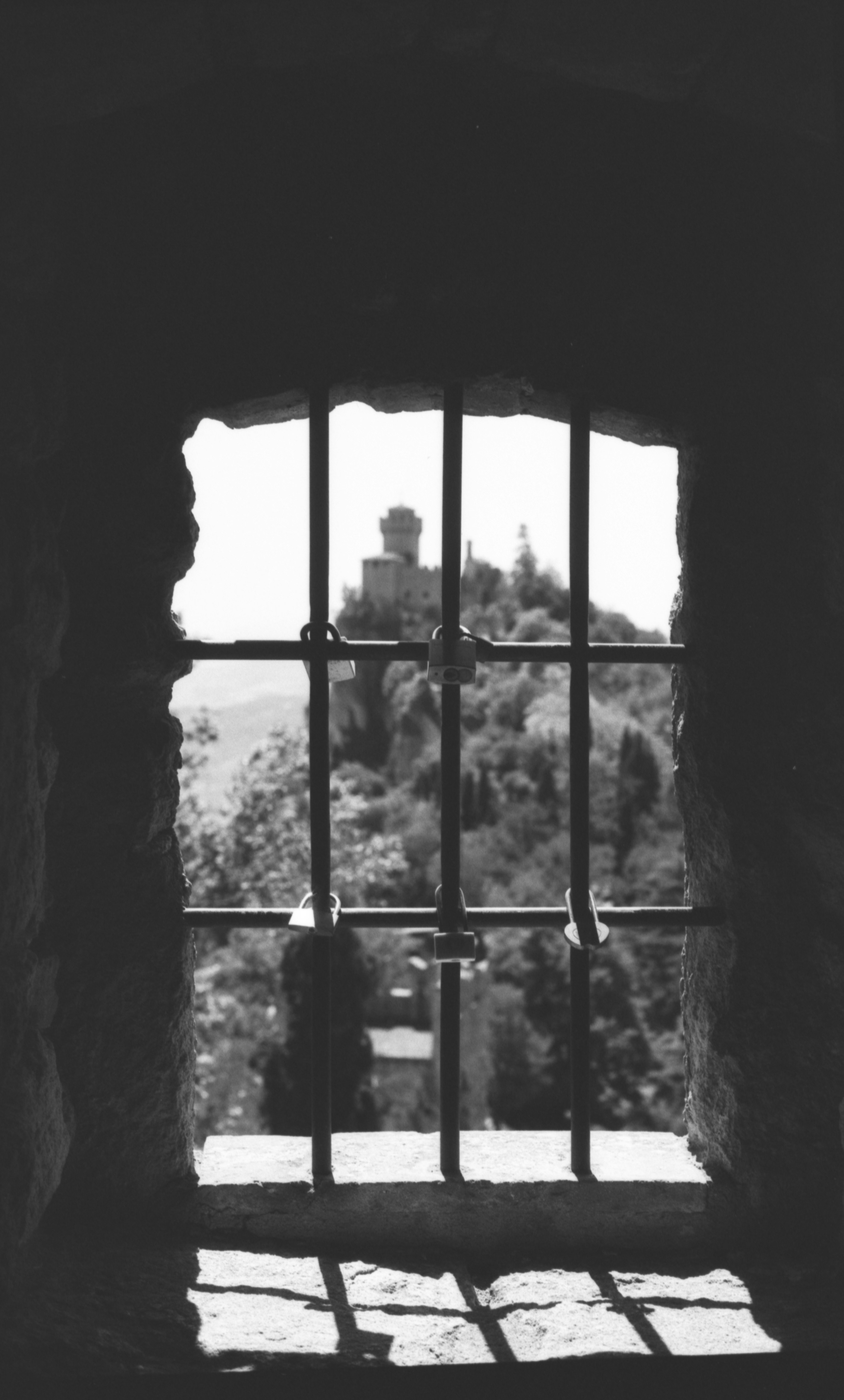 Stone window with vertical and horizontal bars casts shadows onto a sunlit sill, revealing a distant landscape beyond the grate.