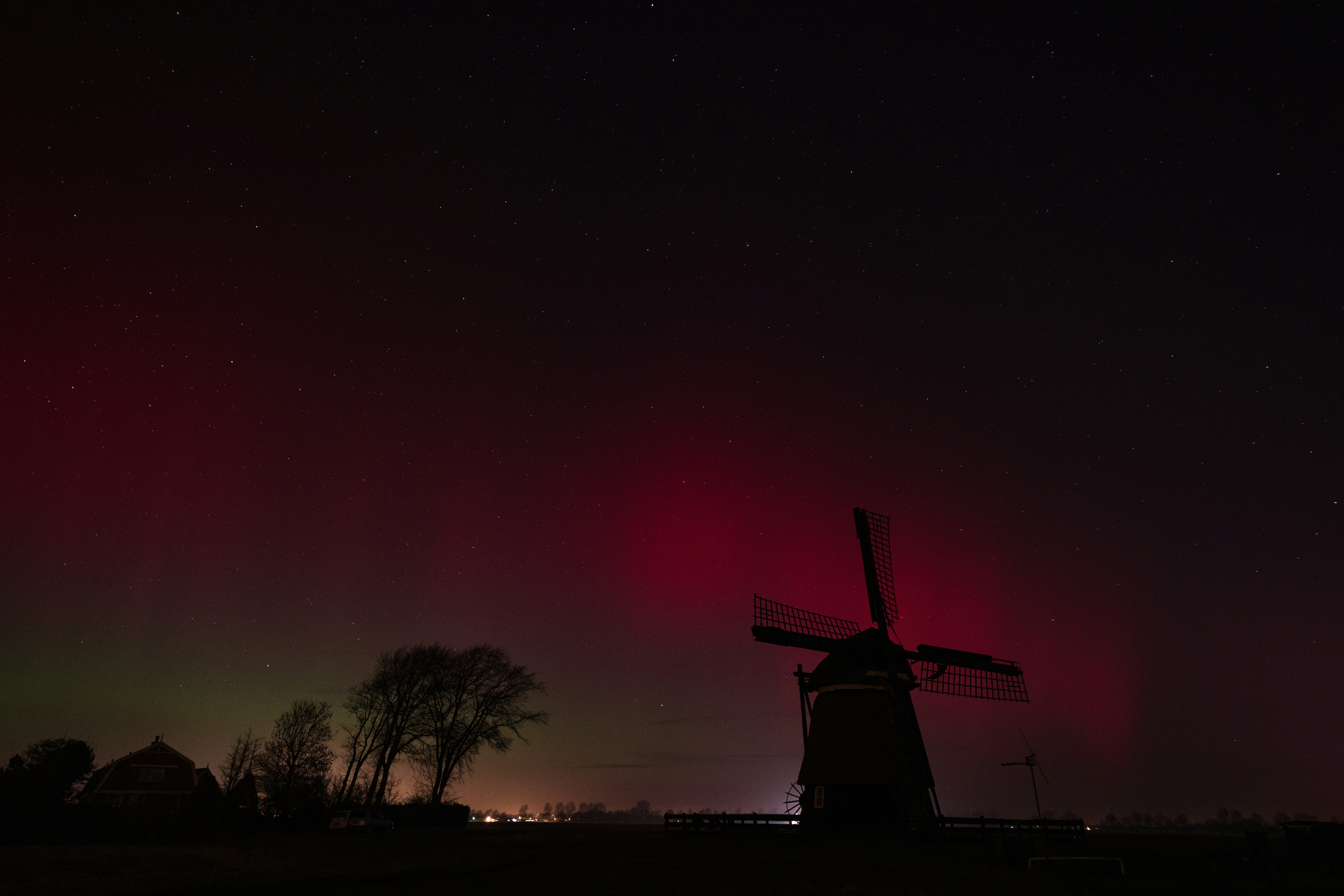 Aurora-like red and green hues sweep above a solitary windmill and rural silhouettes against a starry night sky.