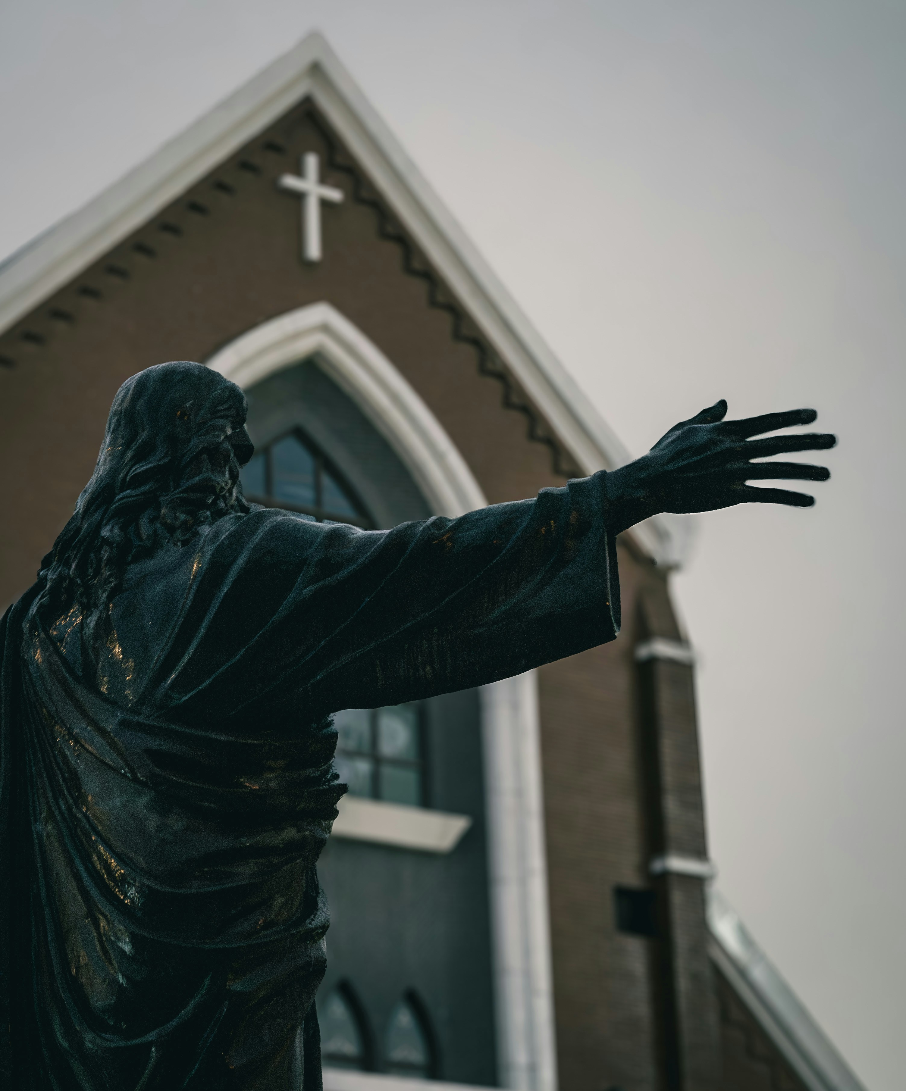 Bronze statue with outstretched arm in front of a church facade, emphasizing gesture against a pale sky.