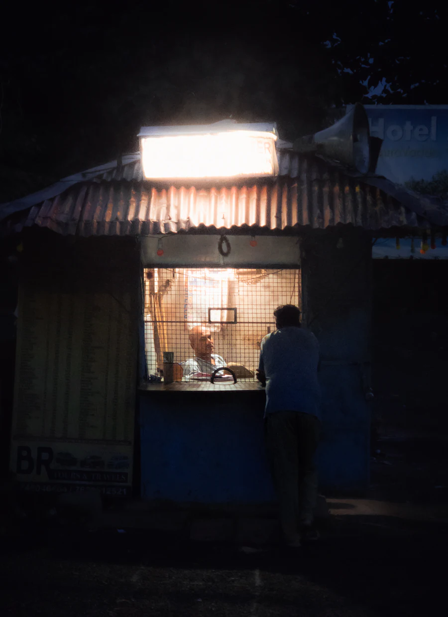 A small roadside snack kiosk glows warmly from inside, with a vendor behind a mesh window and a customer outside at dusk. The scene emphasizes the intimate, dimly lit interaction at a street-side stall; the image captures a logo-style counter look and a soft, warm ambience.