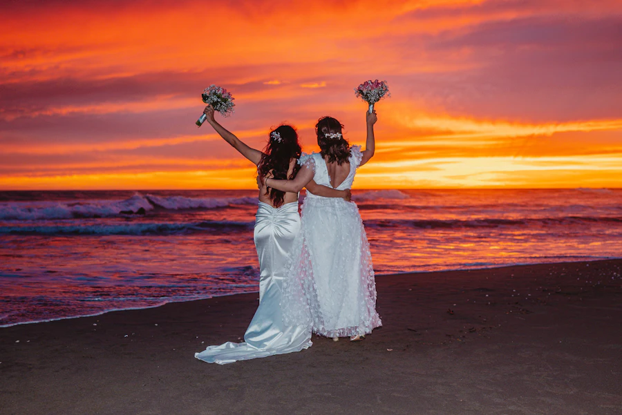 Two brides in white gowns stand arm in arm at the shore during a vivid sunset, holding bouquets as waves roll in the background.