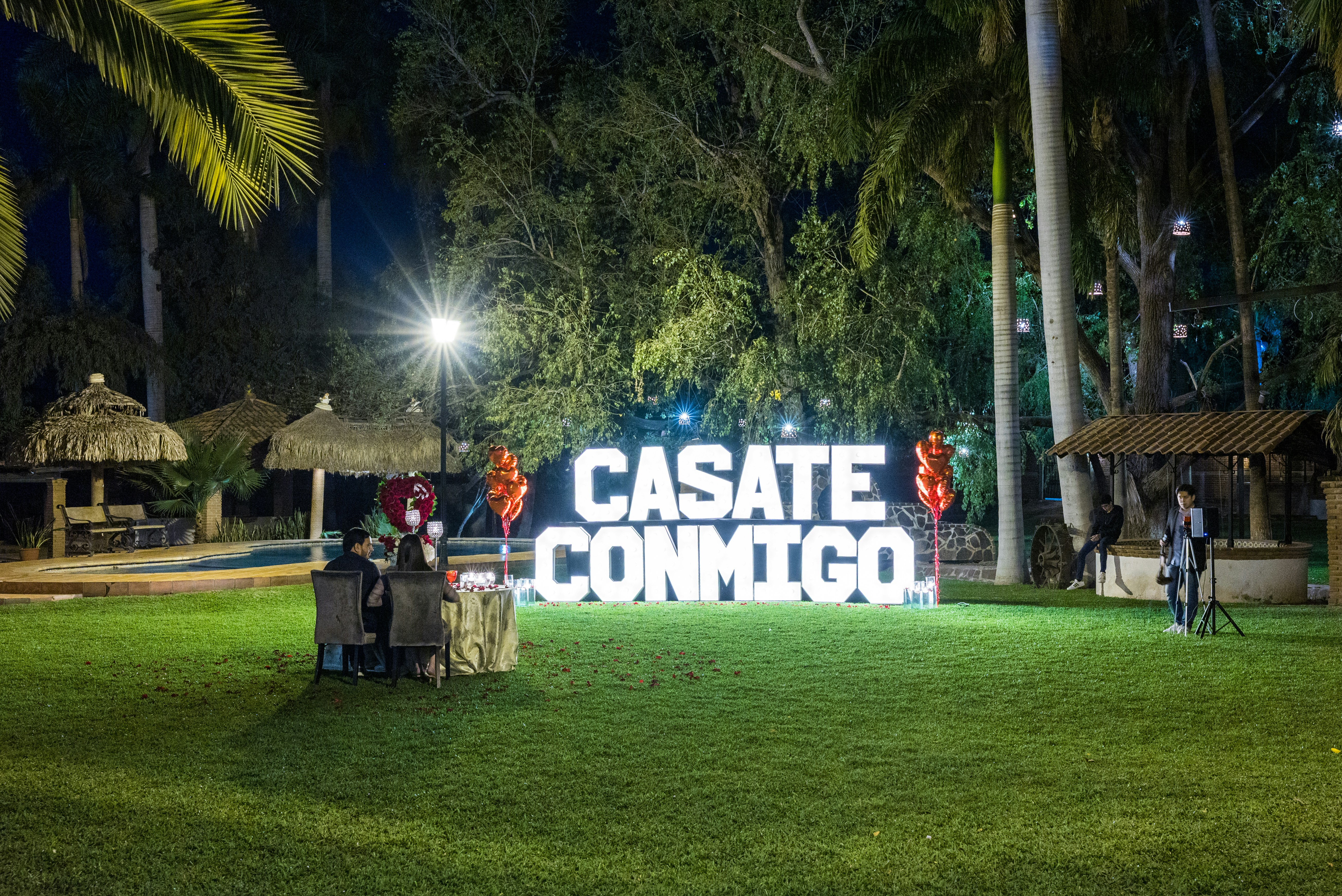 Lit signboard 'CASATE CONMIGO' on a moonlit lawn with audience and palm trees