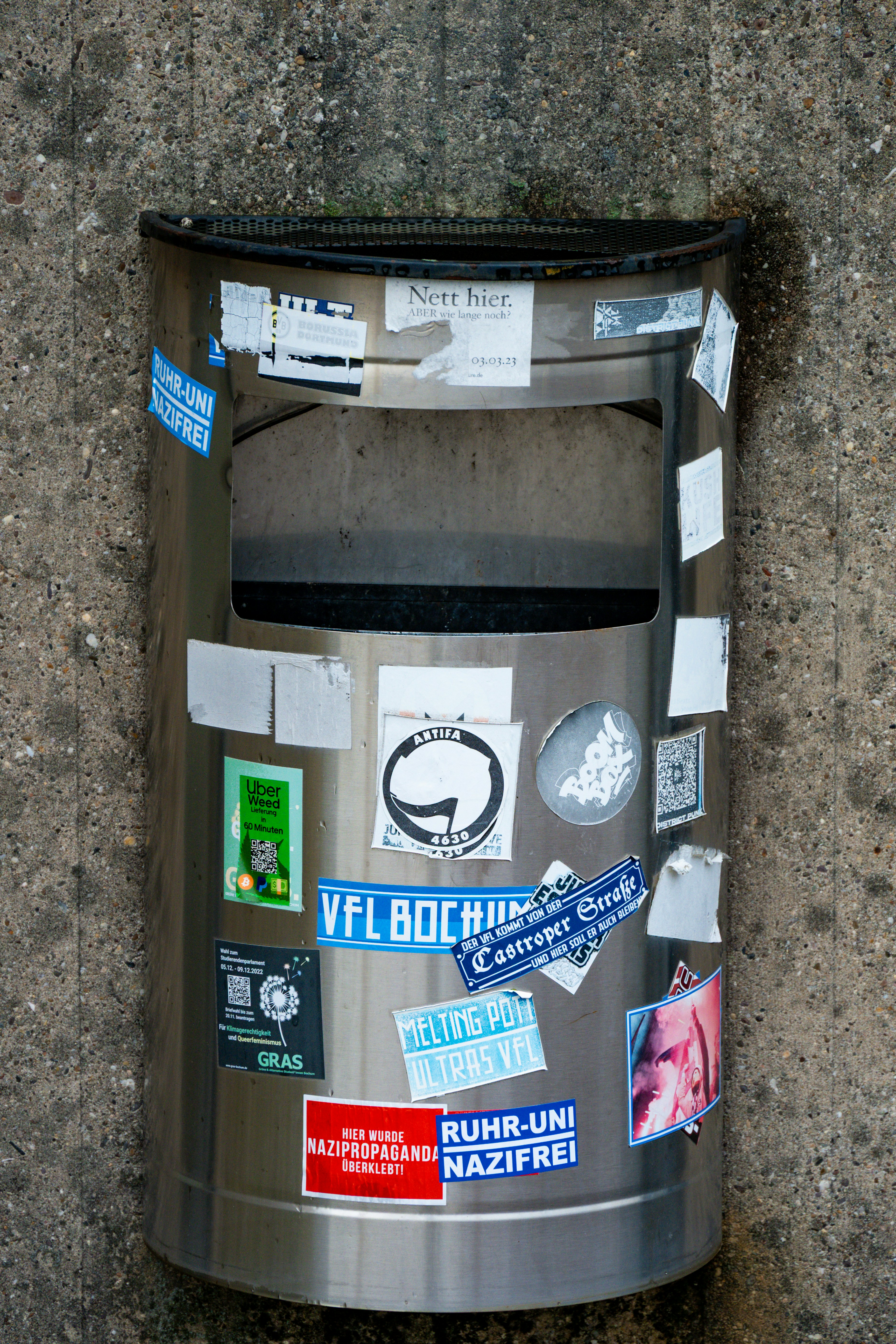 Stickered Newsstand: A Metal Trash Can Wrapped in Posters on a Concrete Wall