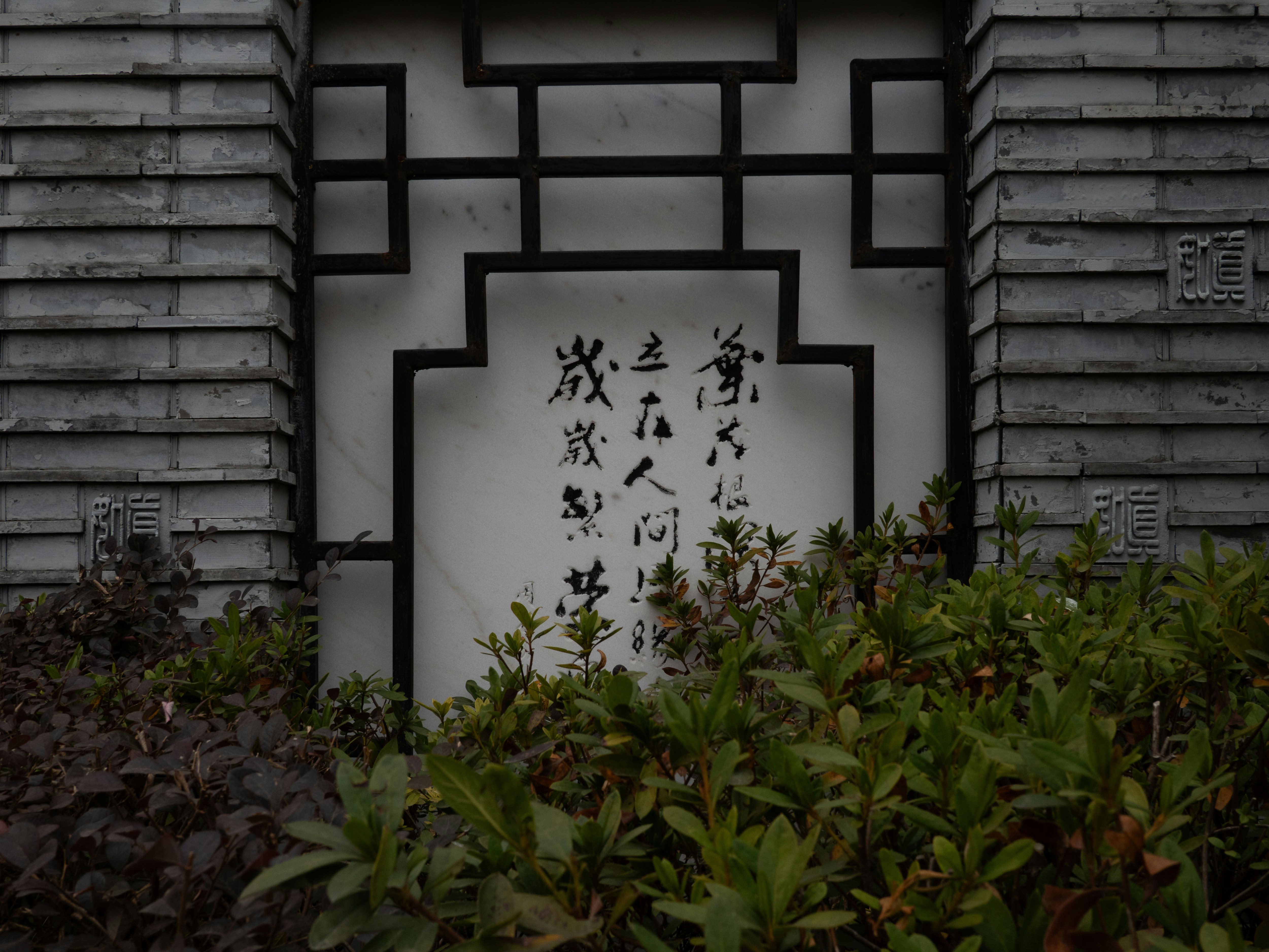 A traditional stone wall with a decorative panel and geometric frame sits behind overgrown shrubs; the scene emphasizes a carved panel with vertical script as the focal point.