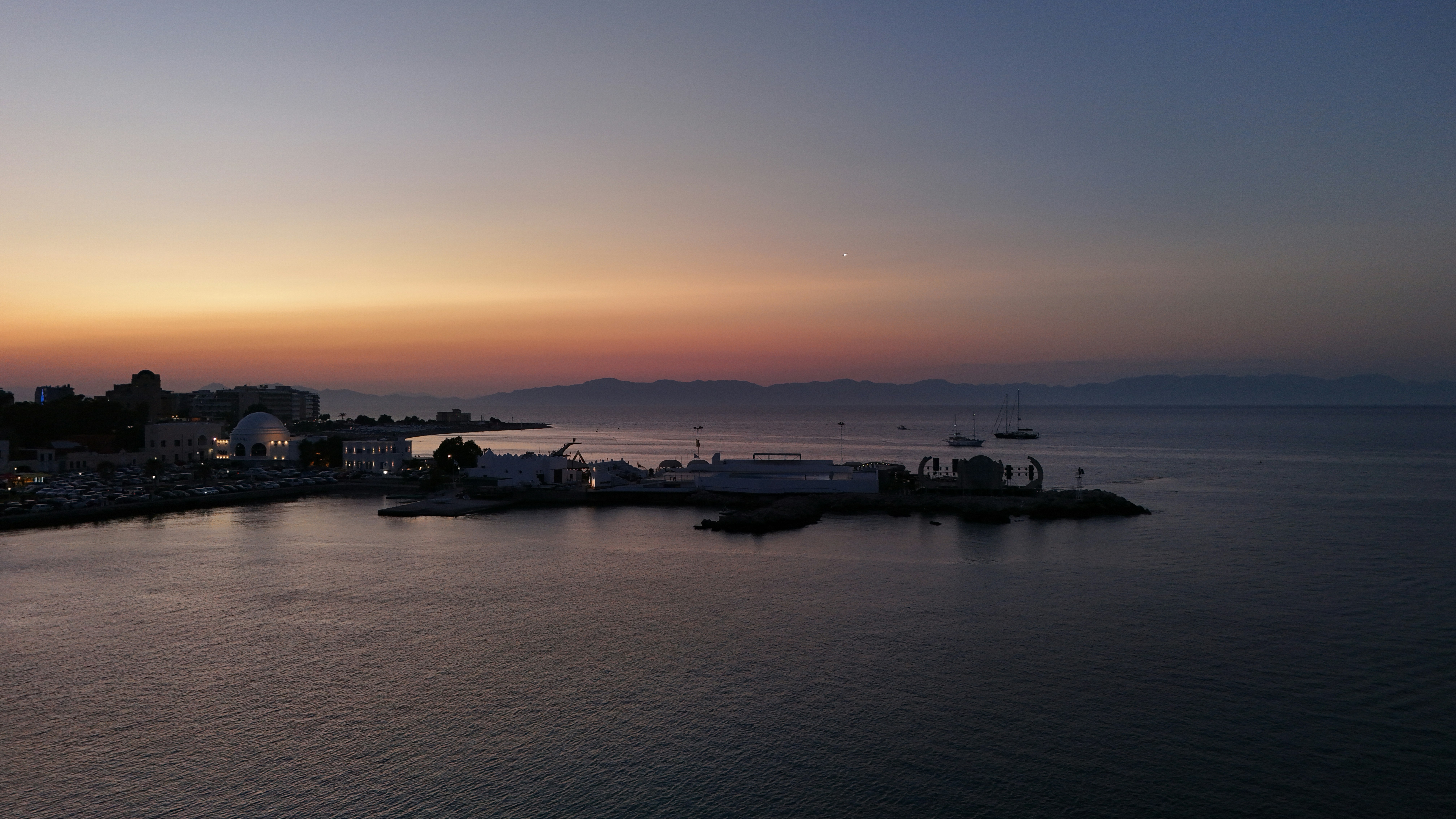 Coastal city skyline at dusk with calm ocean water.