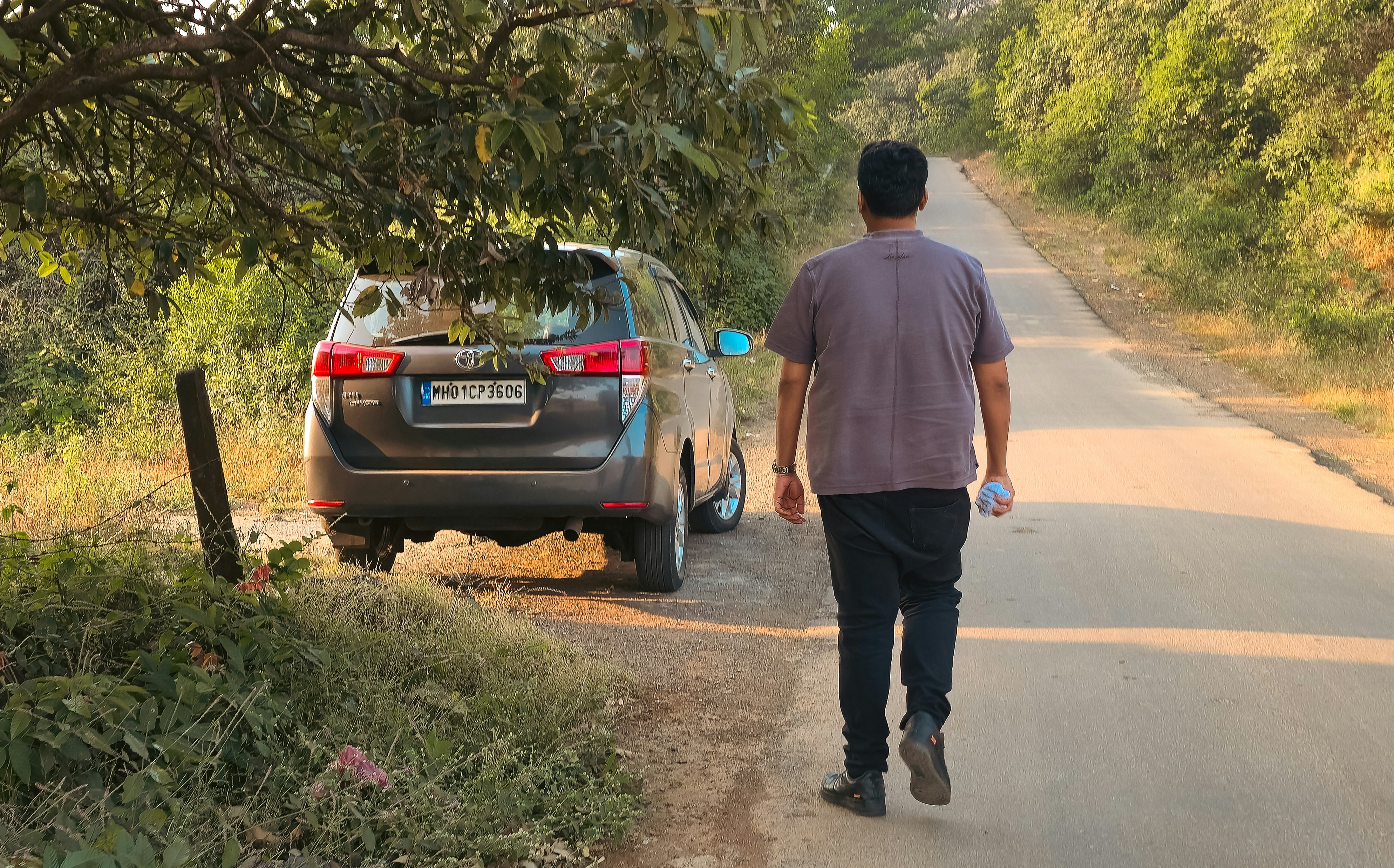 A man walks away along a rural road with a parked SUV to the left; natural light bathes the scene and foliage frames the path. This is a candid street-shot, not a logo, illustration, painting, or cartoon.