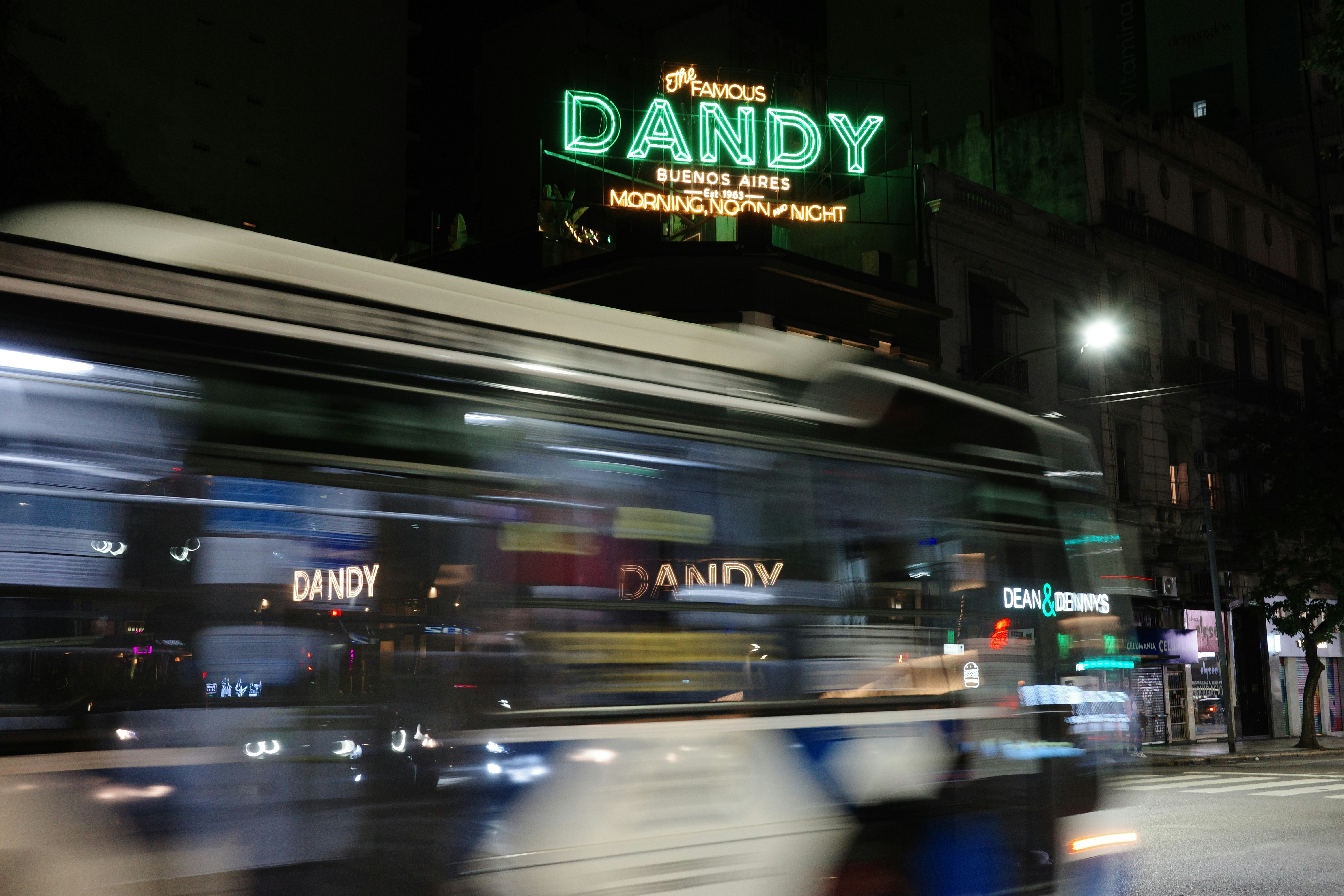 Motion through neon nights: a blurred bus crossfade past DANDY neon